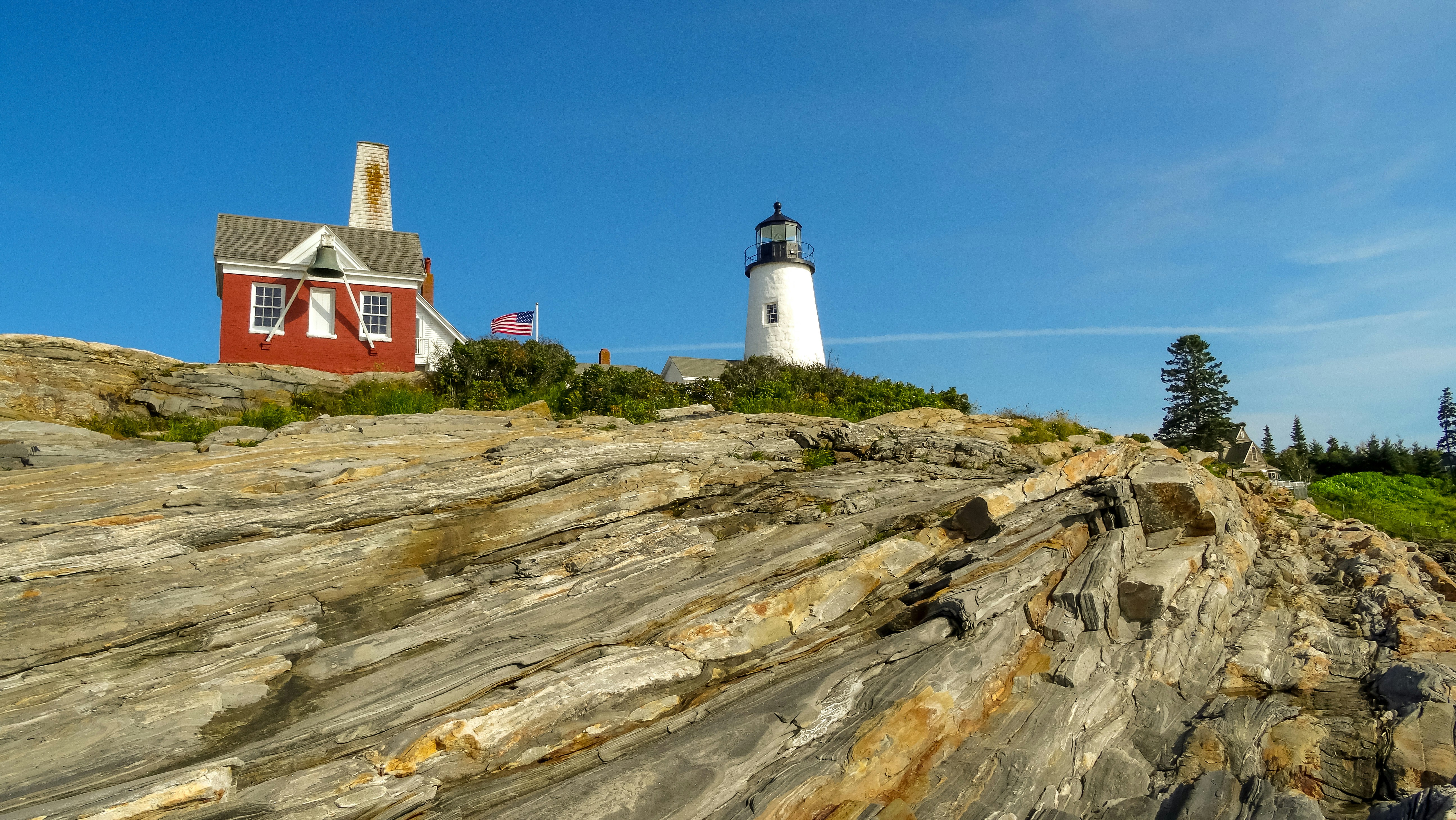 Historic lighthouse perched on rugged rocks, flanked by a charming red building and surrounded by lush greenery.