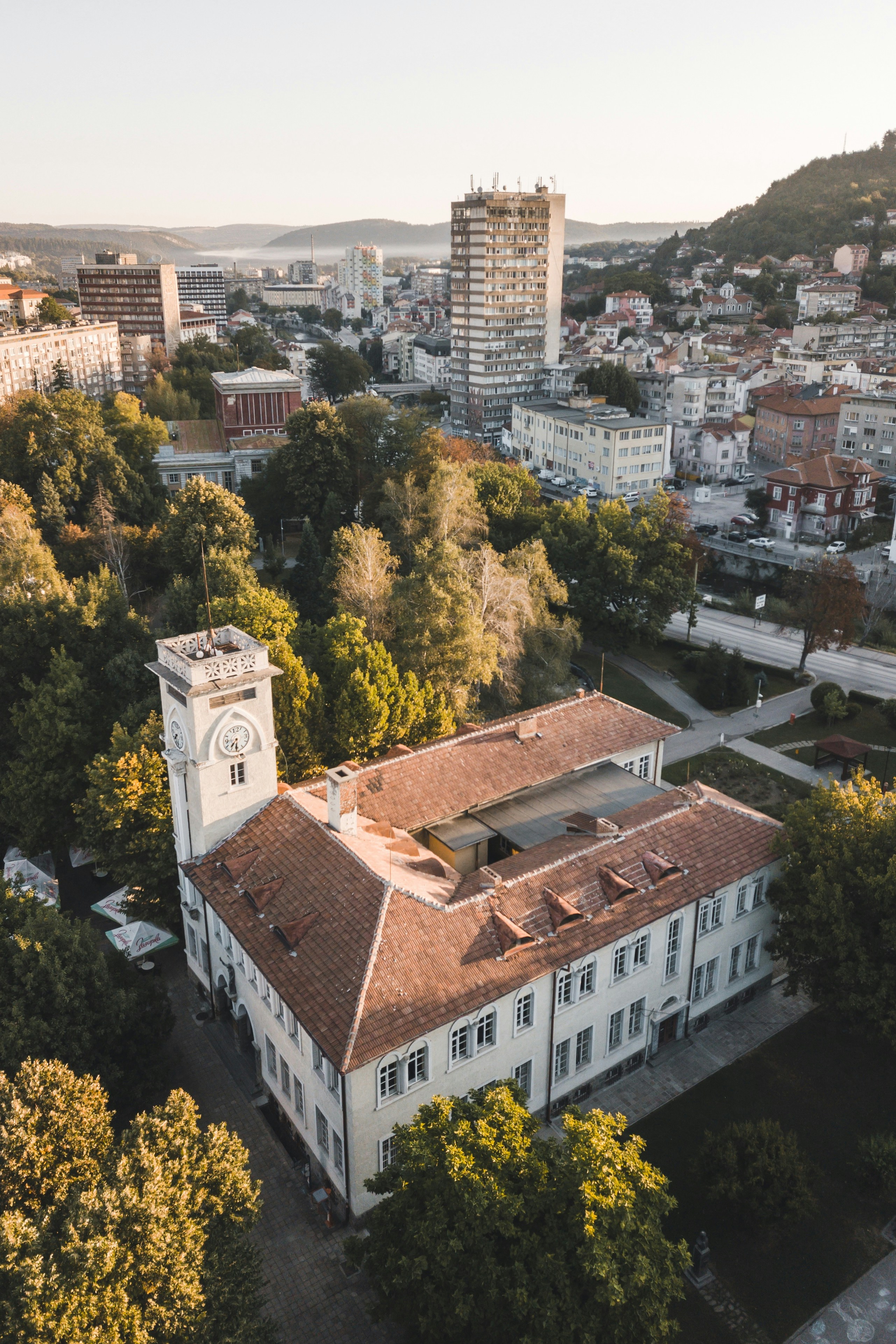 Aerial view showcasing a historical building with a clock tower amidst a vibrant urban landscape, surrounded by lush trees and modern structures.
