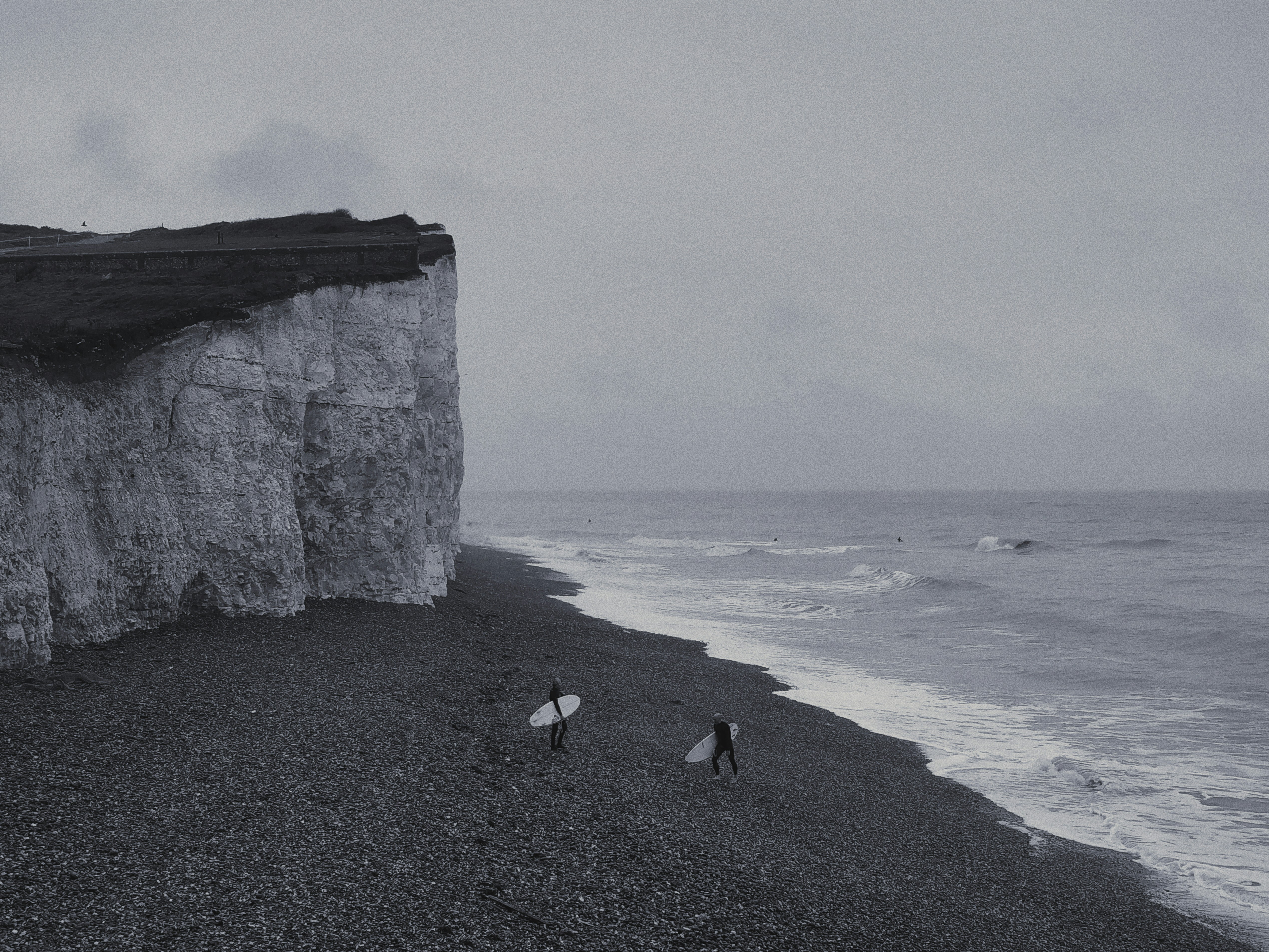 Two surfers walk along a pebbled beach below towering white cliffs under an overcast sky.