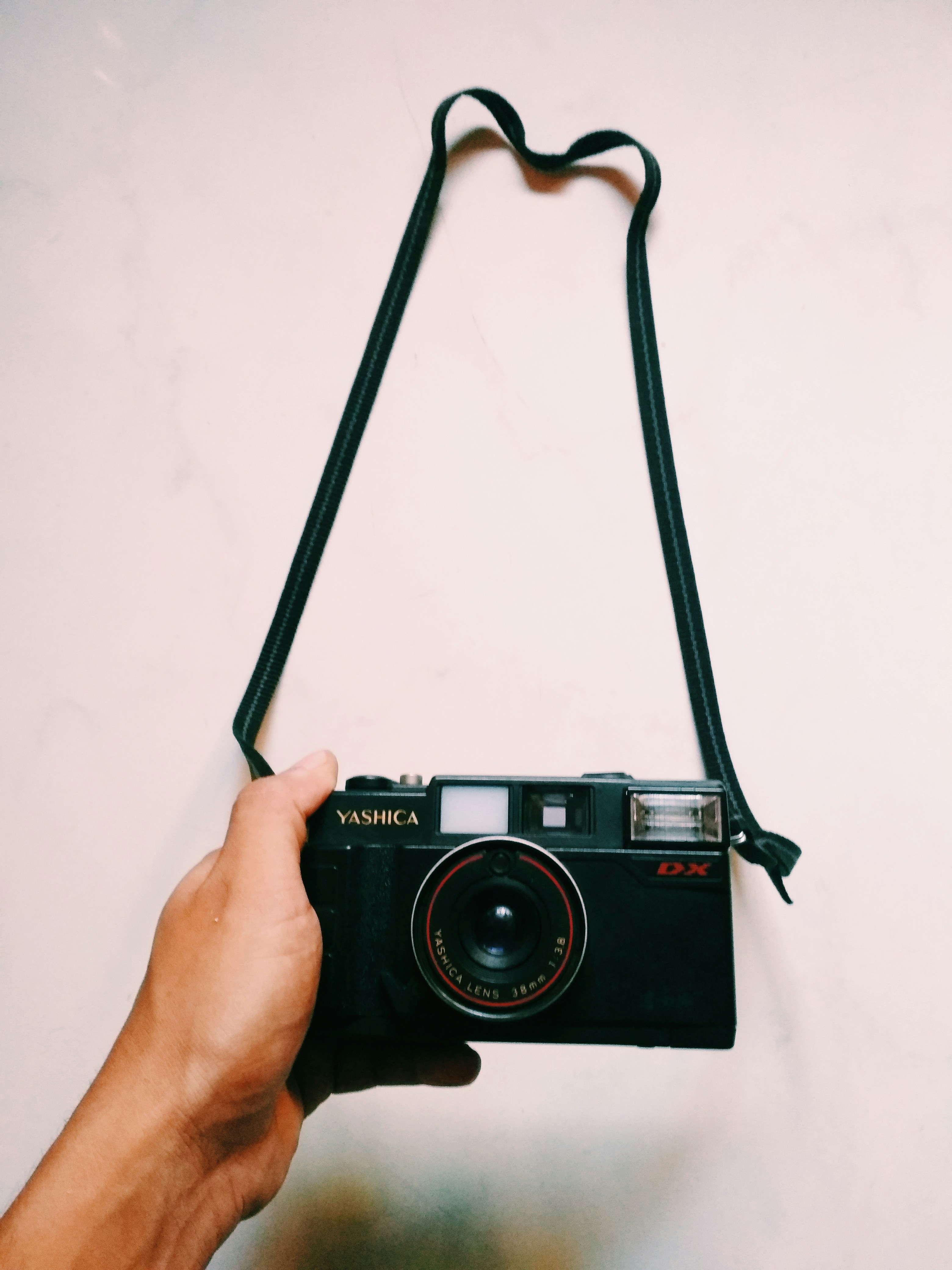 A Yashica DX film camera is held by a hand against a plain pale backdrop, with the lens as the central focal point. The composition emphasizes a vintage device as the primary subject.