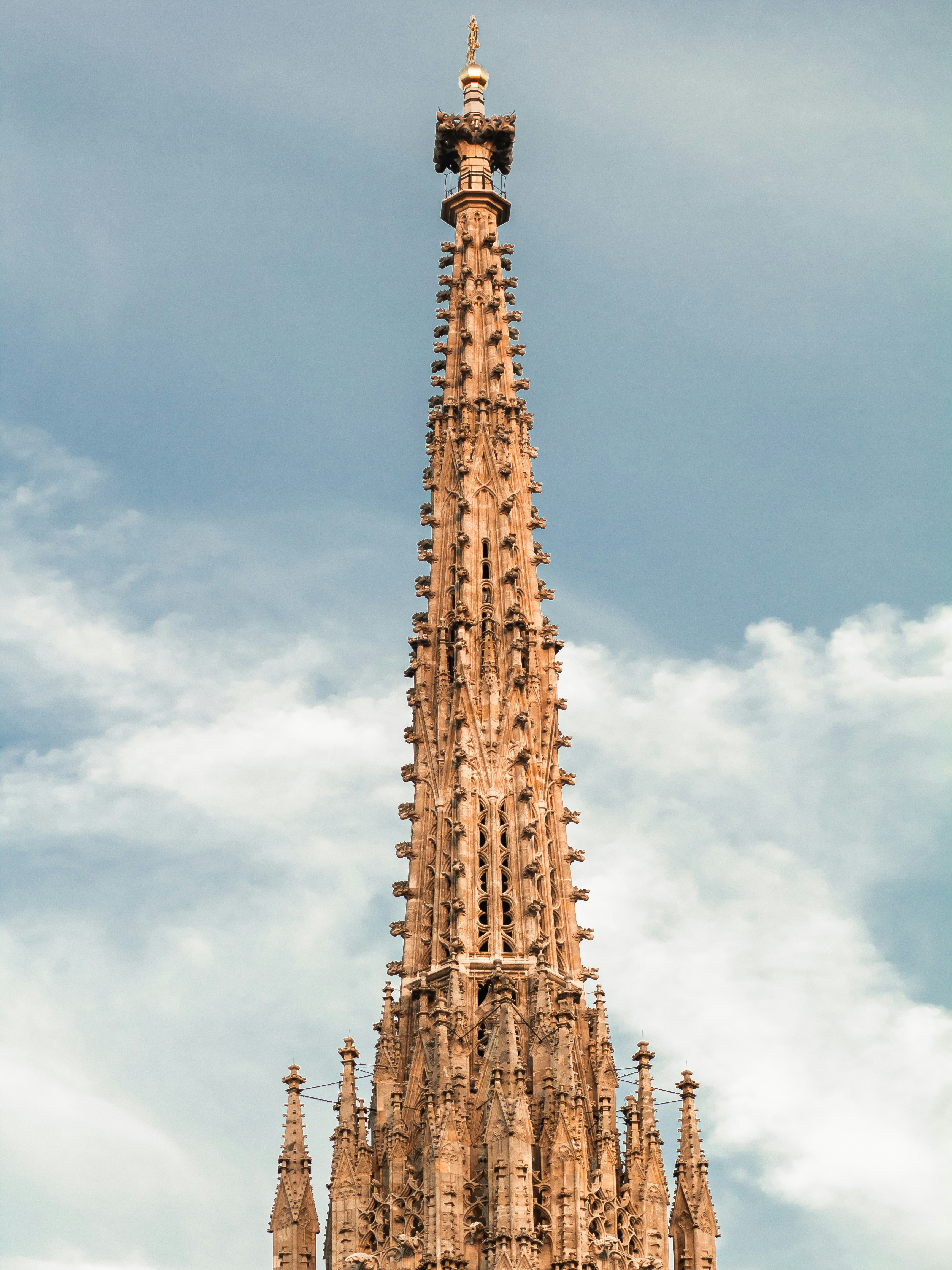 Intricate spire of a Gothic cathedral reaching into the sky, adorned with detailed stonework against a backdrop of soft clouds.