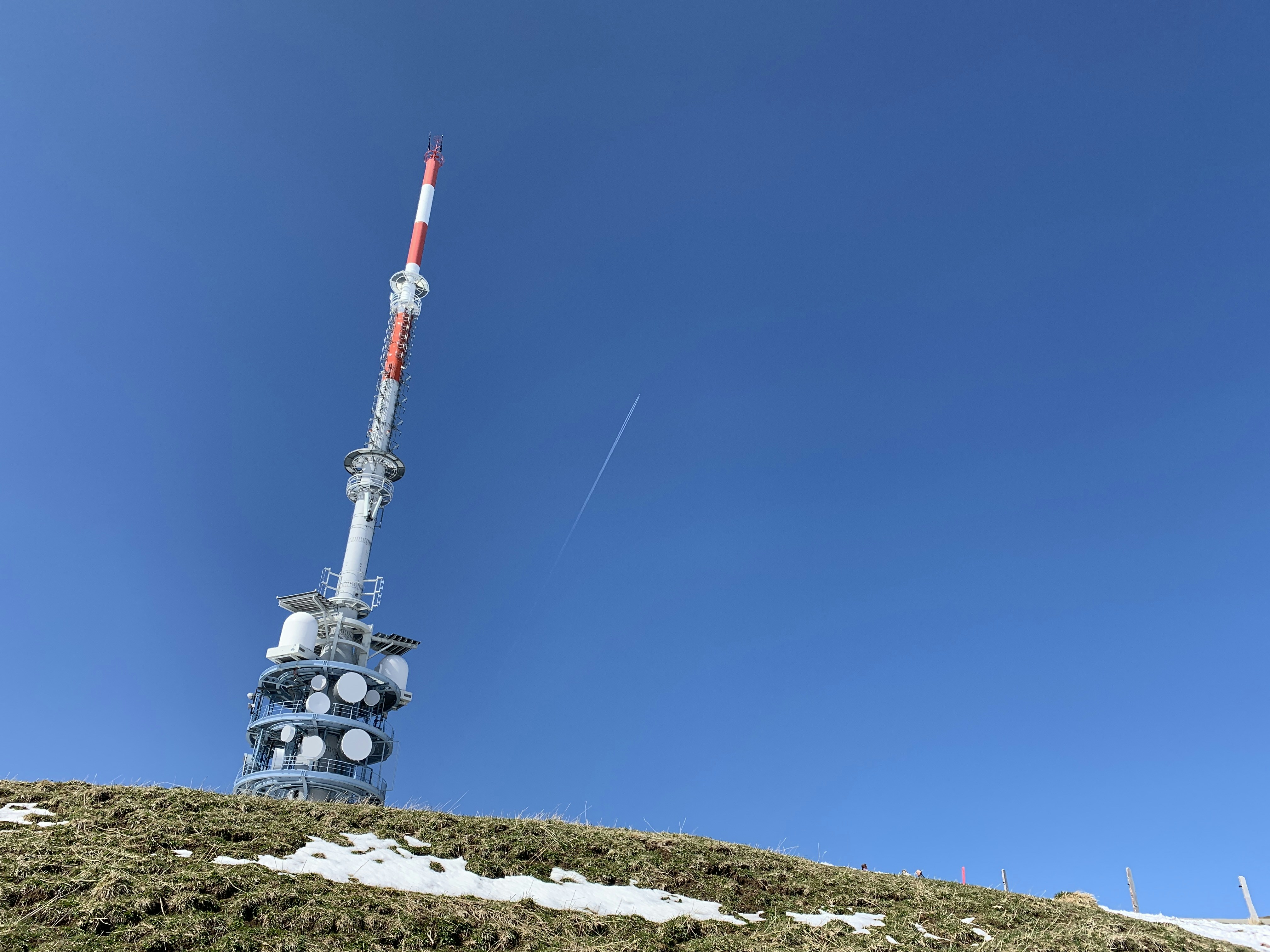 Communications tower rises against a clear blue sky, surrounded by patches of snow and grass. A contrail streaks across the background.