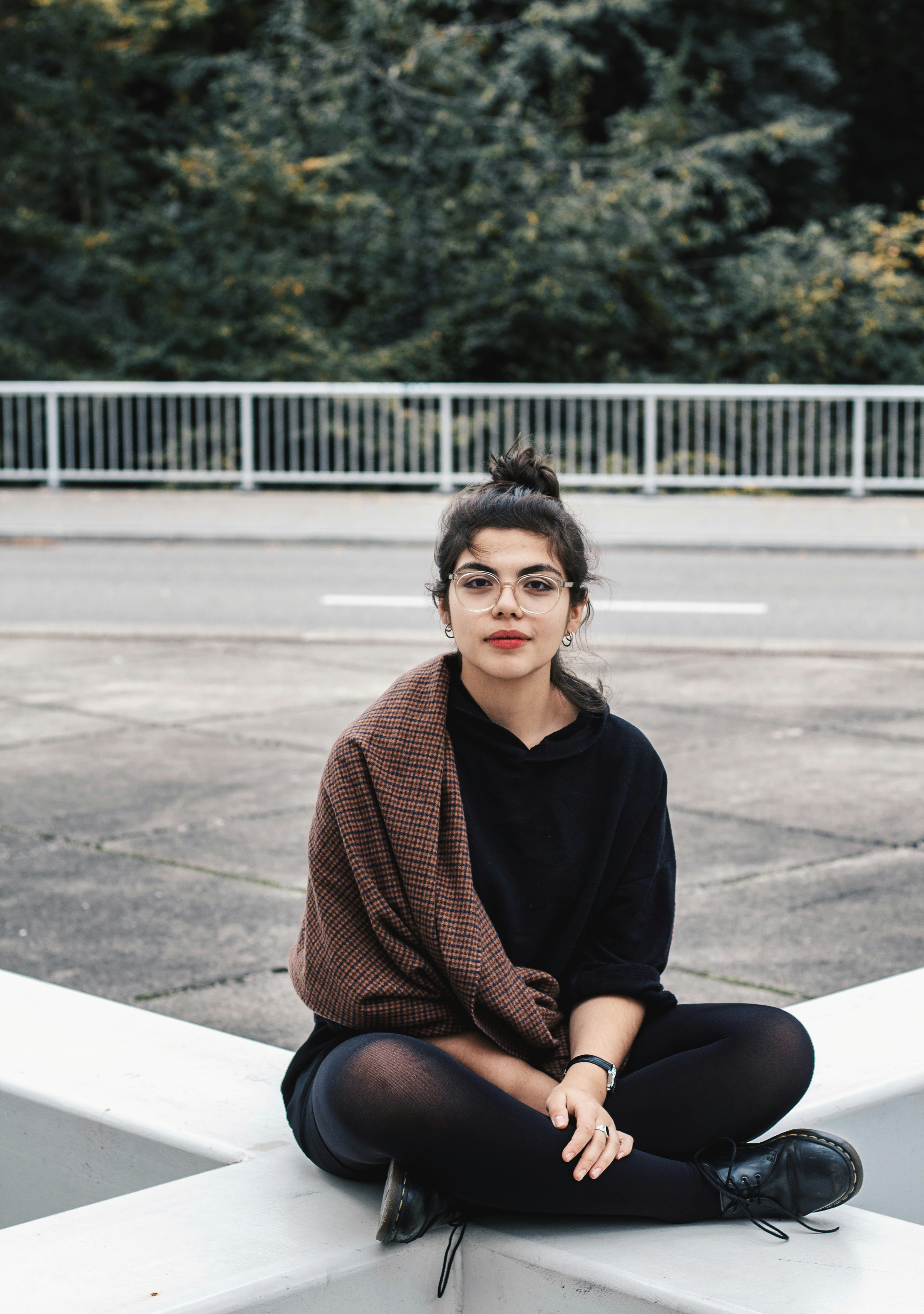Young woman seated cross-legged on a concrete structure, wearing a black outfit and a patterned shawl, with a blurred urban backdrop.