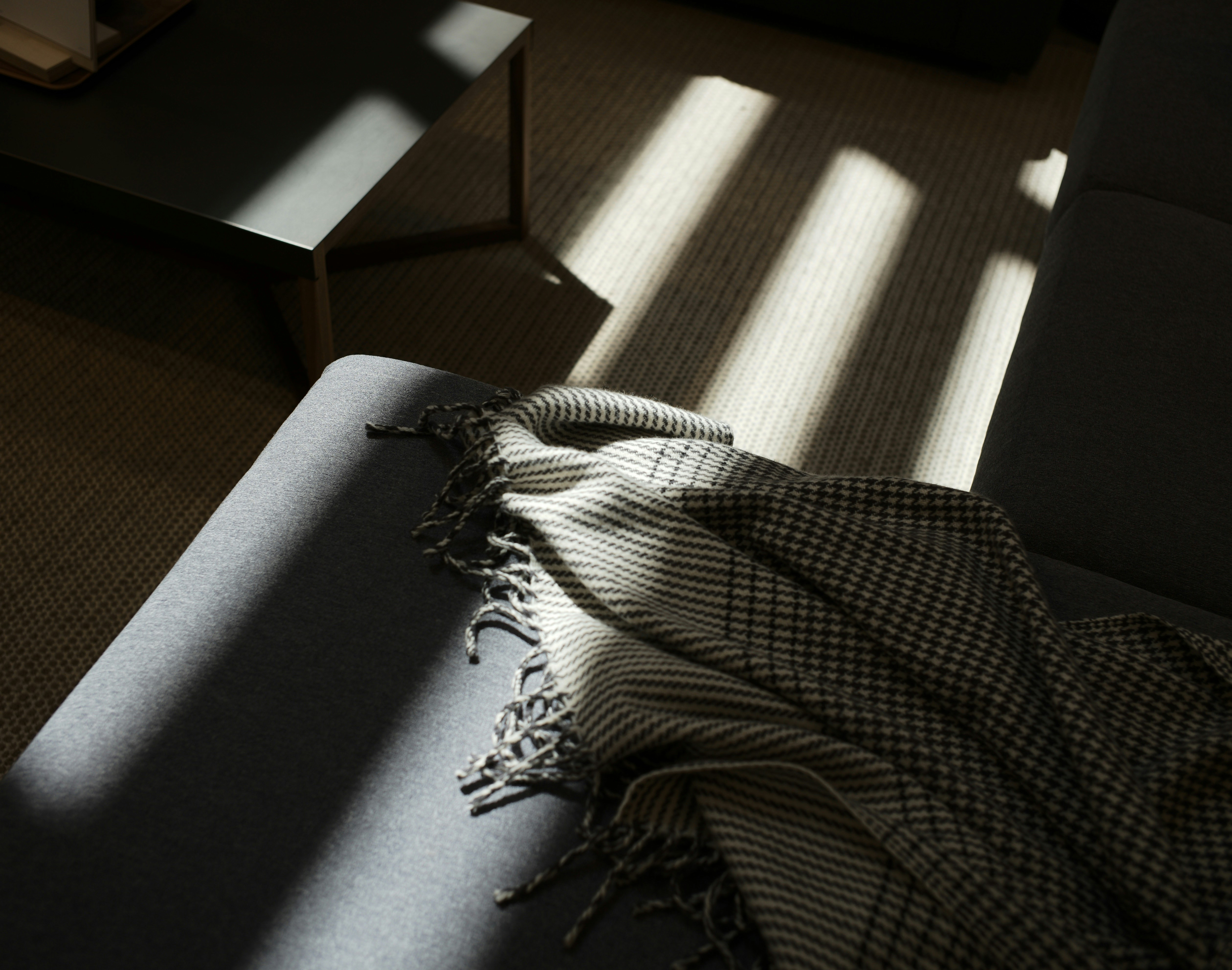 Sunbeams slice through a living room, casting striped light across a sofa. A fringed throw drapes the foreground, highlighting texture and a tranquil mood.