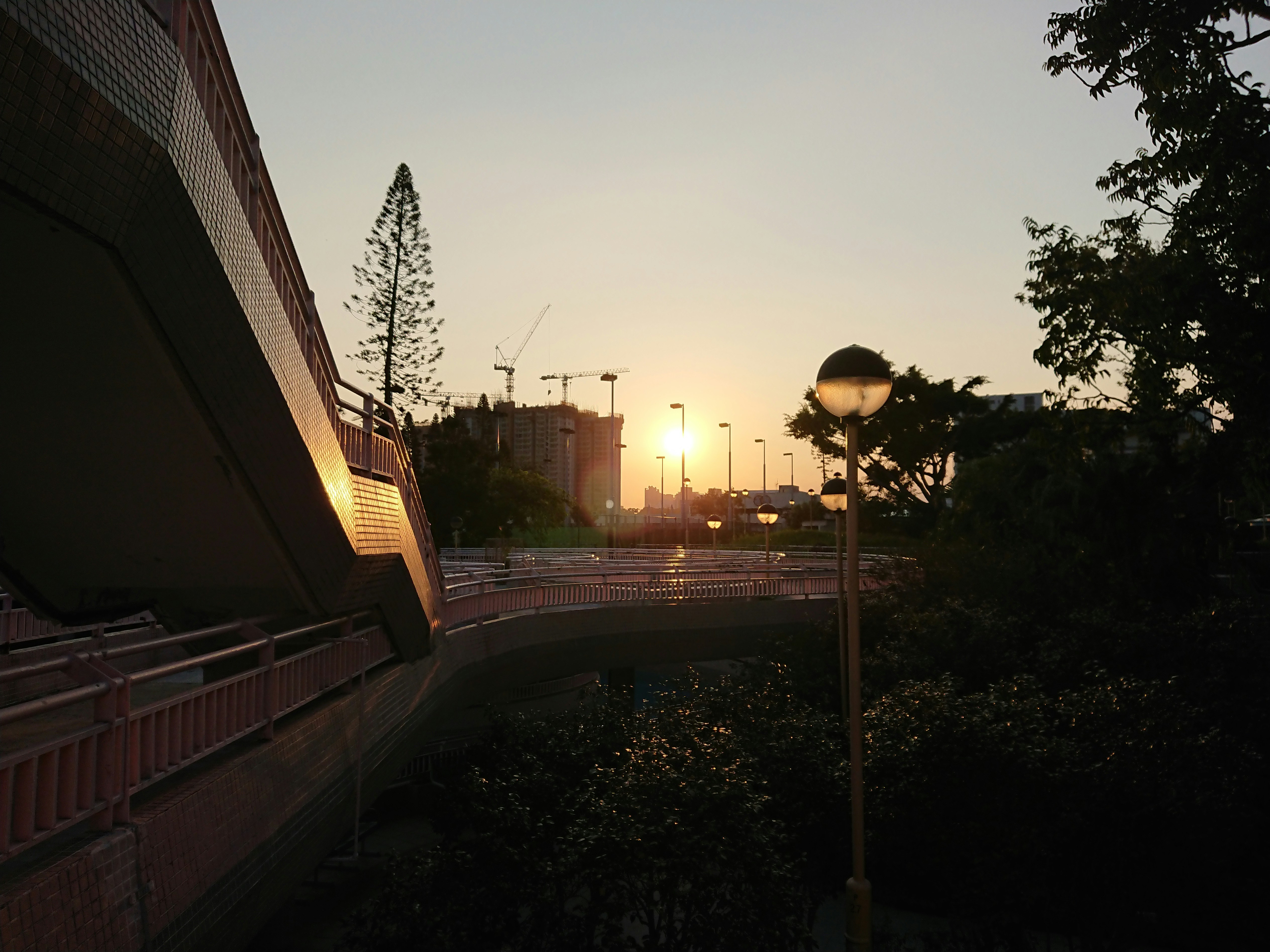 Golden hour bathes a curved pedestrian bridge with warm light and a line of lampposts; silhouettes of buildings frame the horizon. Water reflects the sunset, adding depth to the composition.