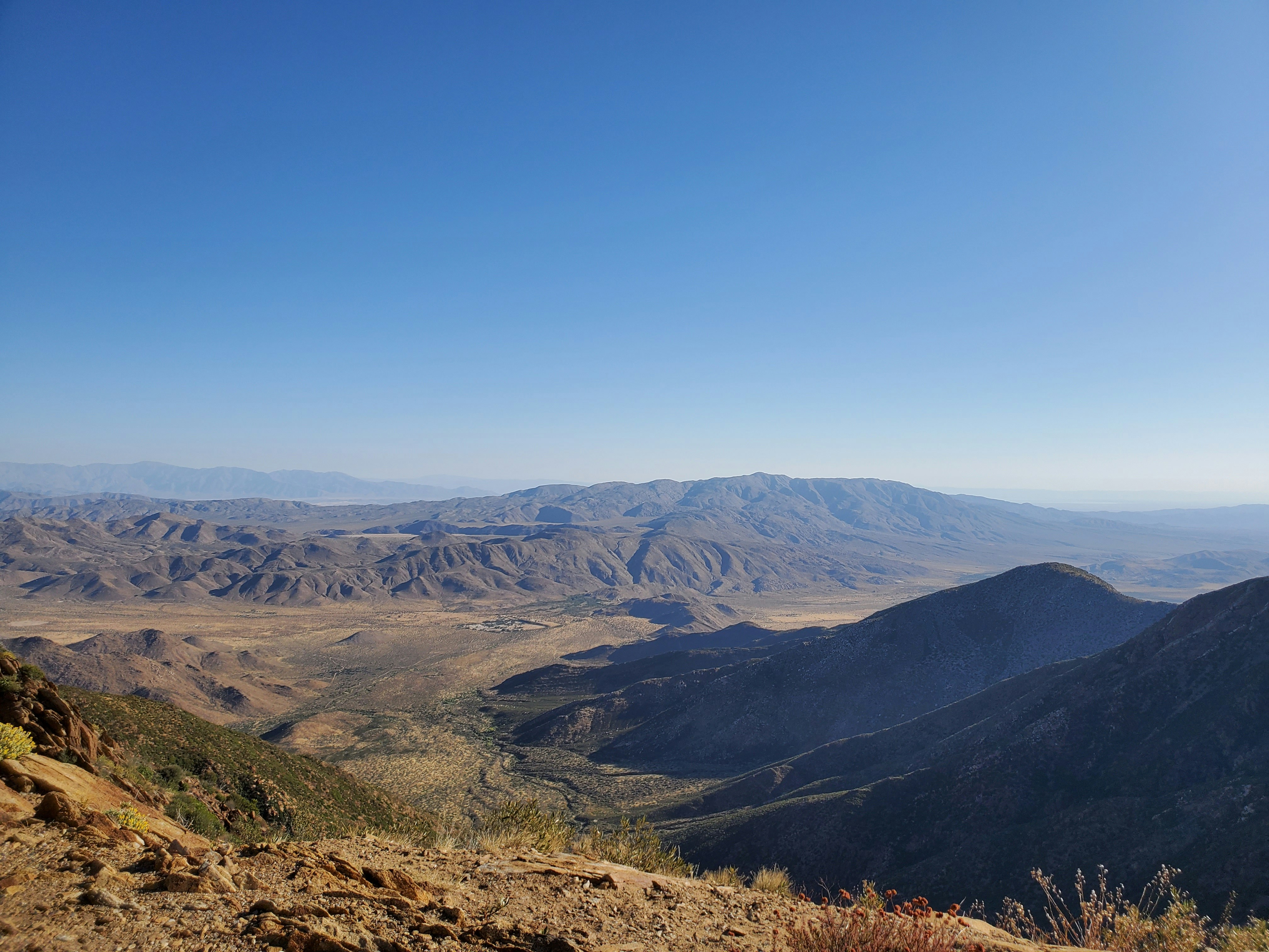 Expansive view of a desert valley beneath a clear blue sky with distant mountain ranges.