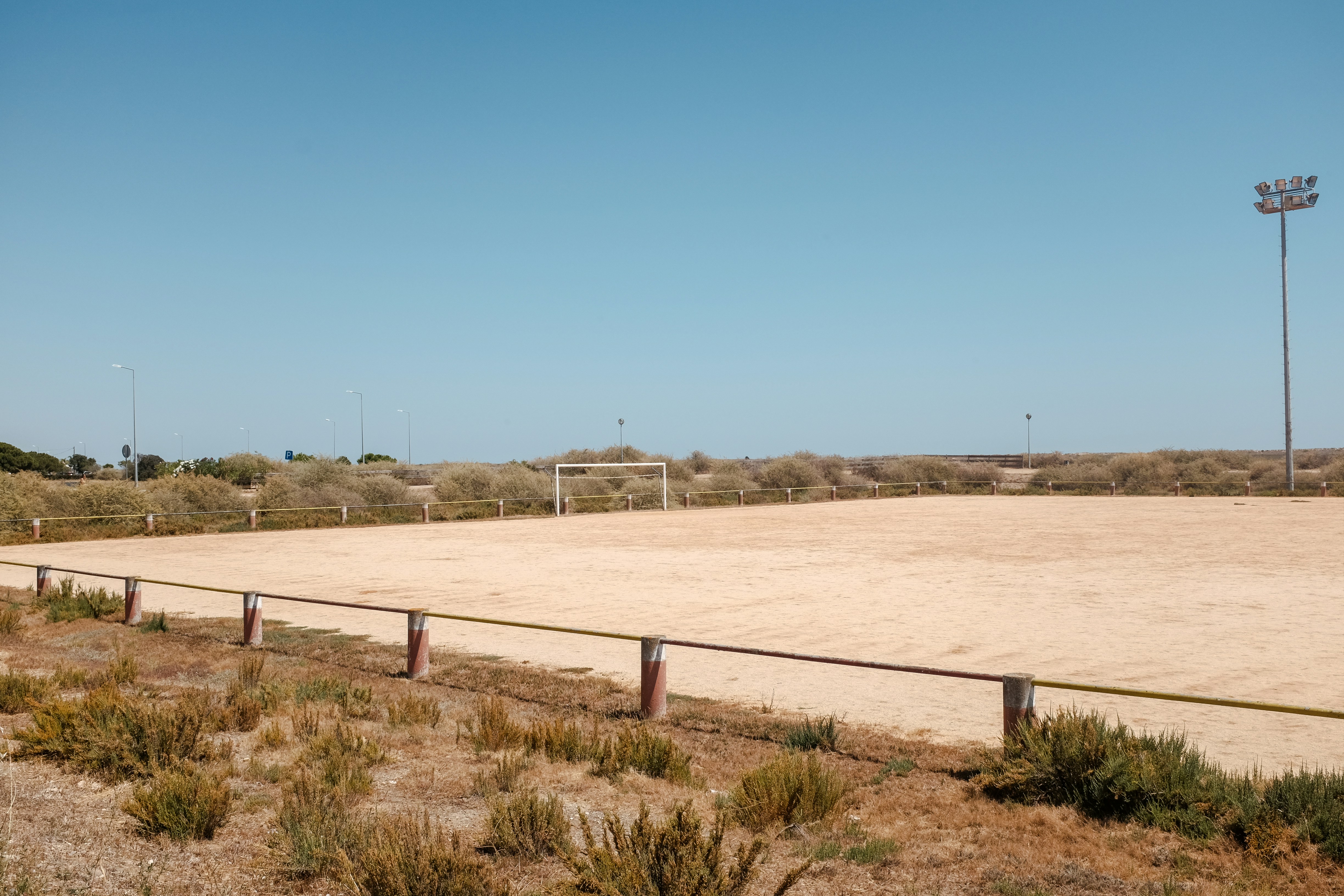 Abandoned soccer field surrounded by sparse vegetation, with goalposts in the distance under a bright blue sky.