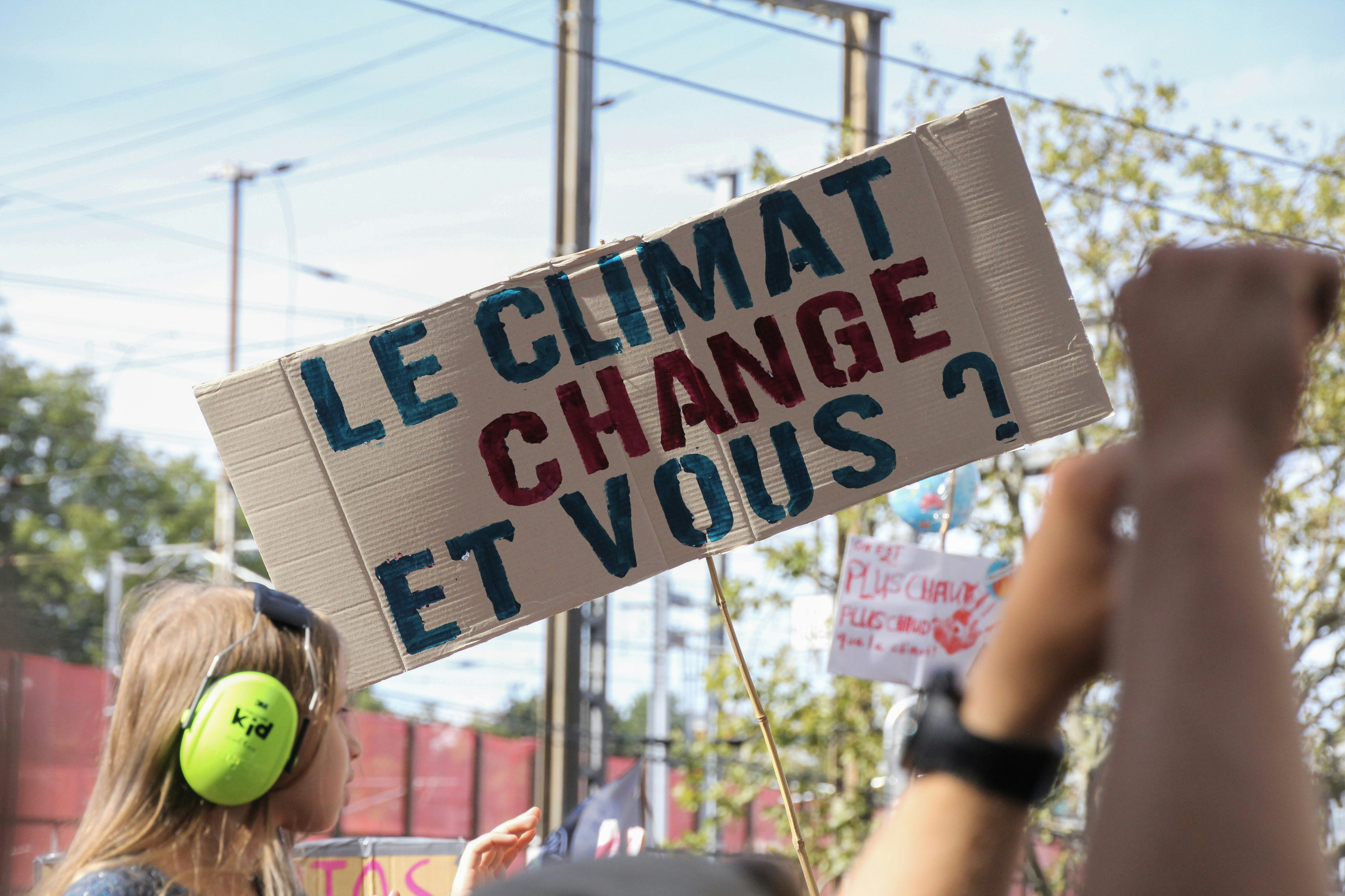 A protestor holds a sign reading 'LE CLIMAT CHANGE ET VOUS?' during a climate demonstration, surrounded by fellow activists. The atmosphere is charged with urgency.