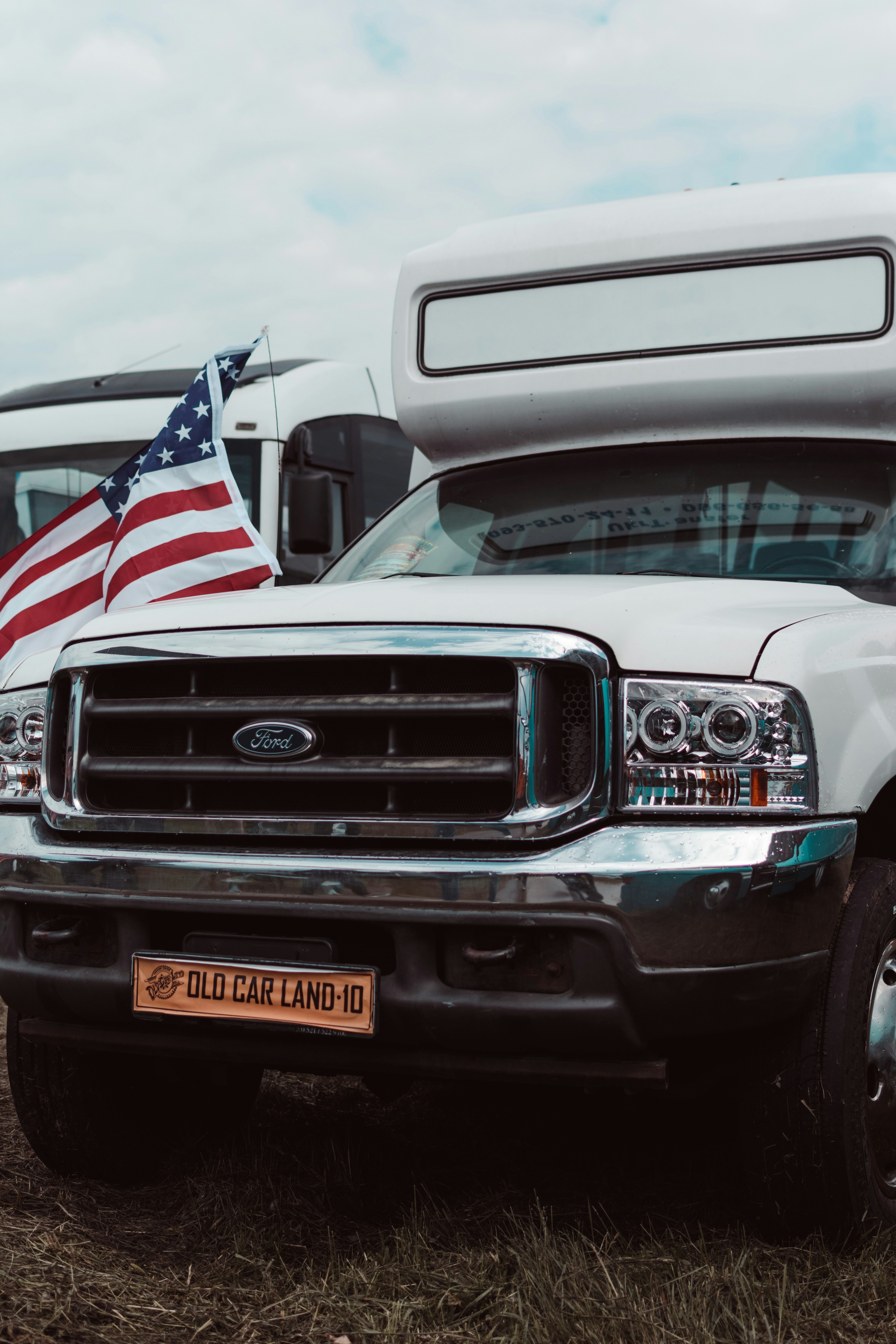 Front view of a Ford truck adorned with American flags, parked amidst a backdrop of other vehicles. The license plate reads 'OLD CAR LAND-10.'