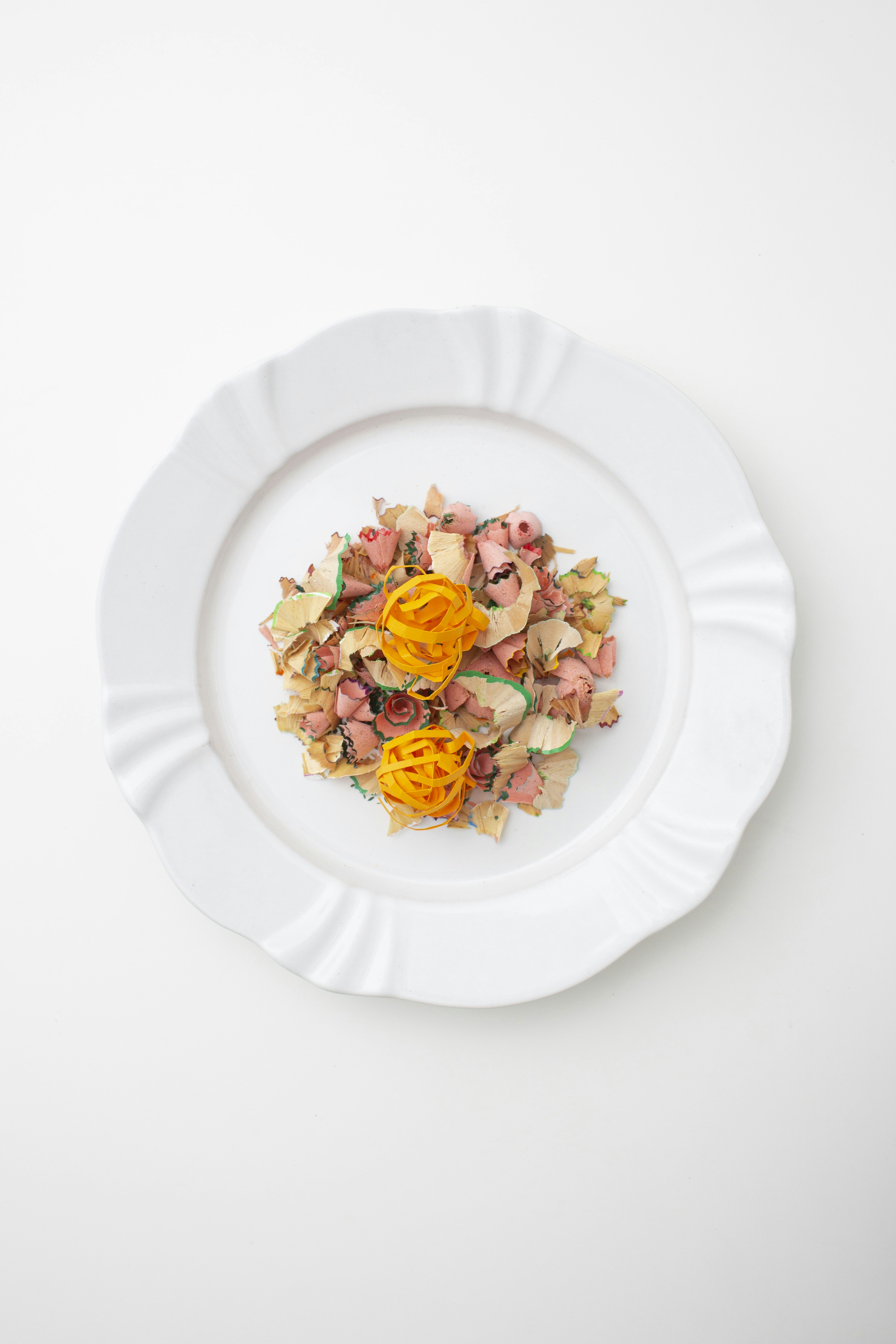 A white plate full of colored pencil chips and curled paper, in a white background | a white plate topped with food on top of a white table