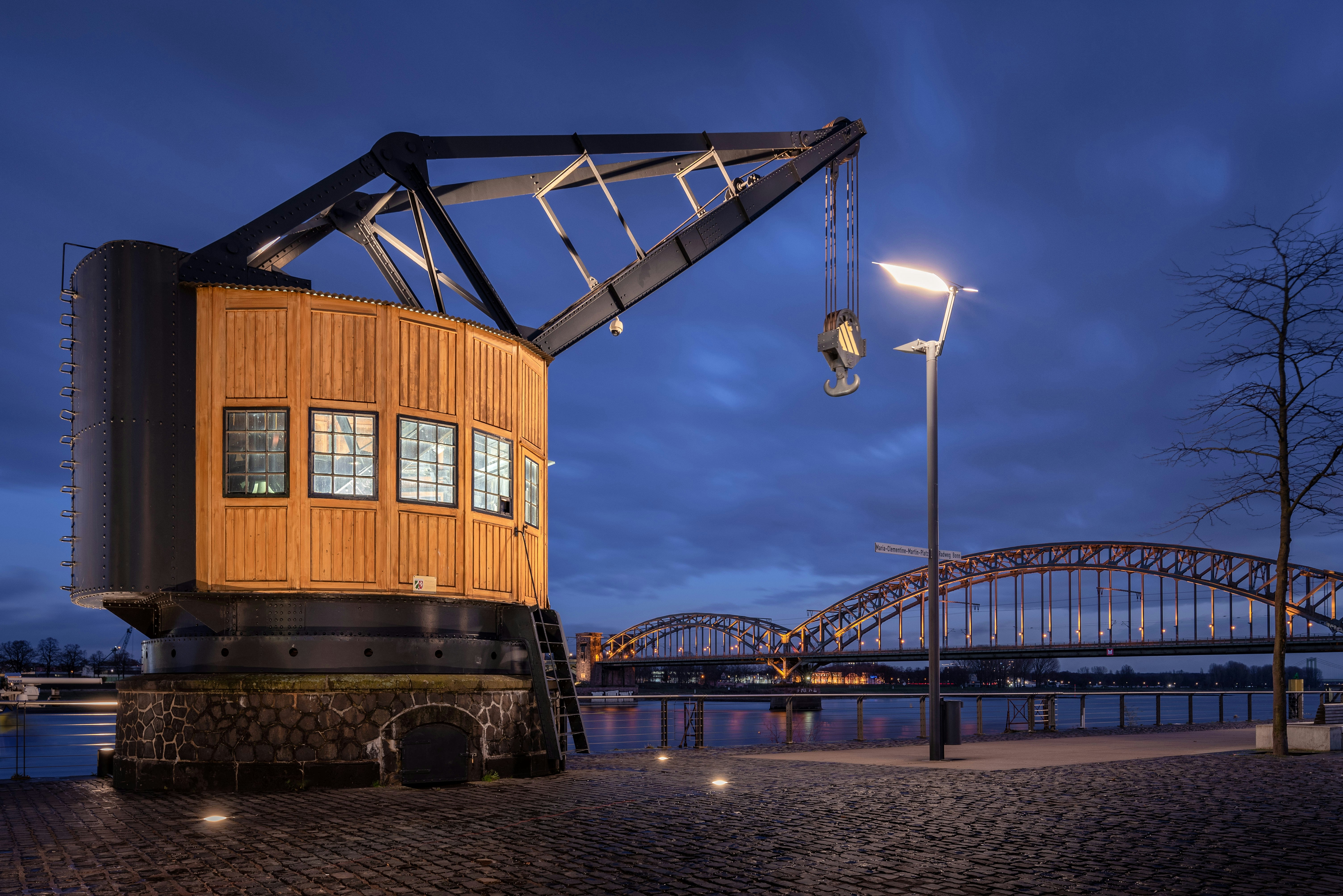 Wooden crane structure illuminated at dusk, with an arched bridge in the background under a deep blue sky.