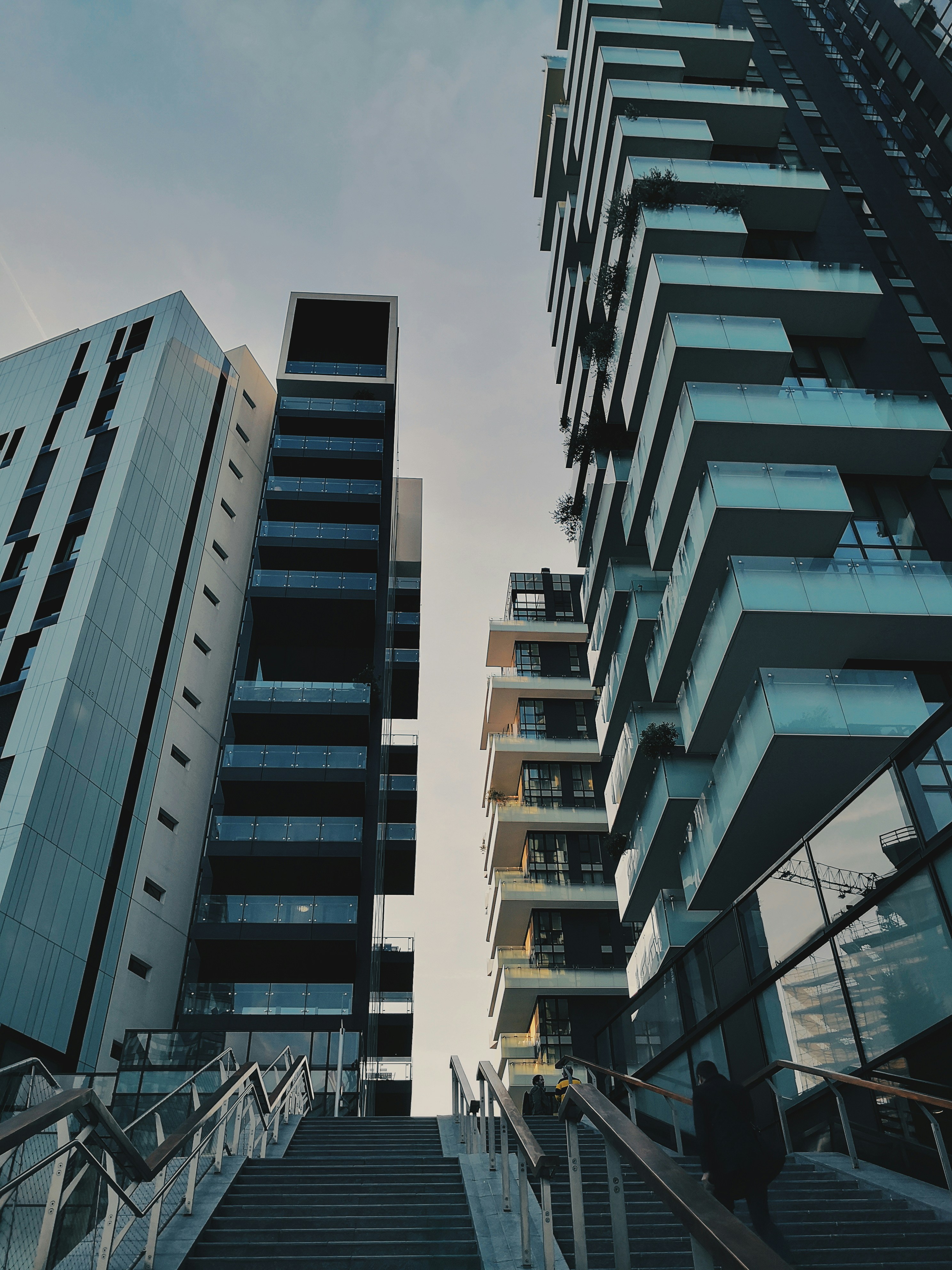 Urban canyon of glass towers flank a central stairway that climbs toward a pale sky, highlighting perspective and architectural geometry.