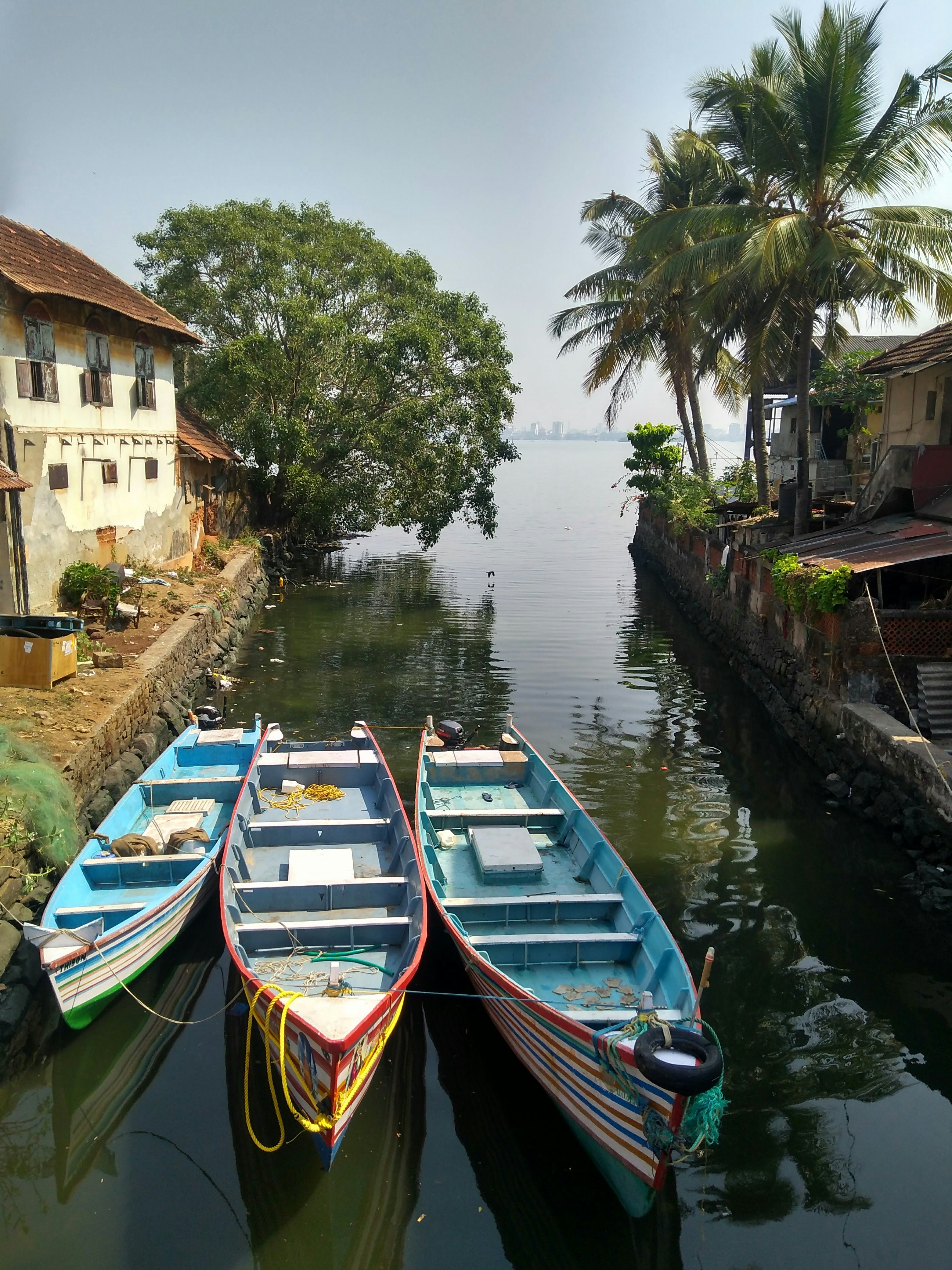 Three colorful fishing boats moored along a serene waterway, flanked by lush greenery and rustic buildings. The calm surface reflects the surrounding scenery.