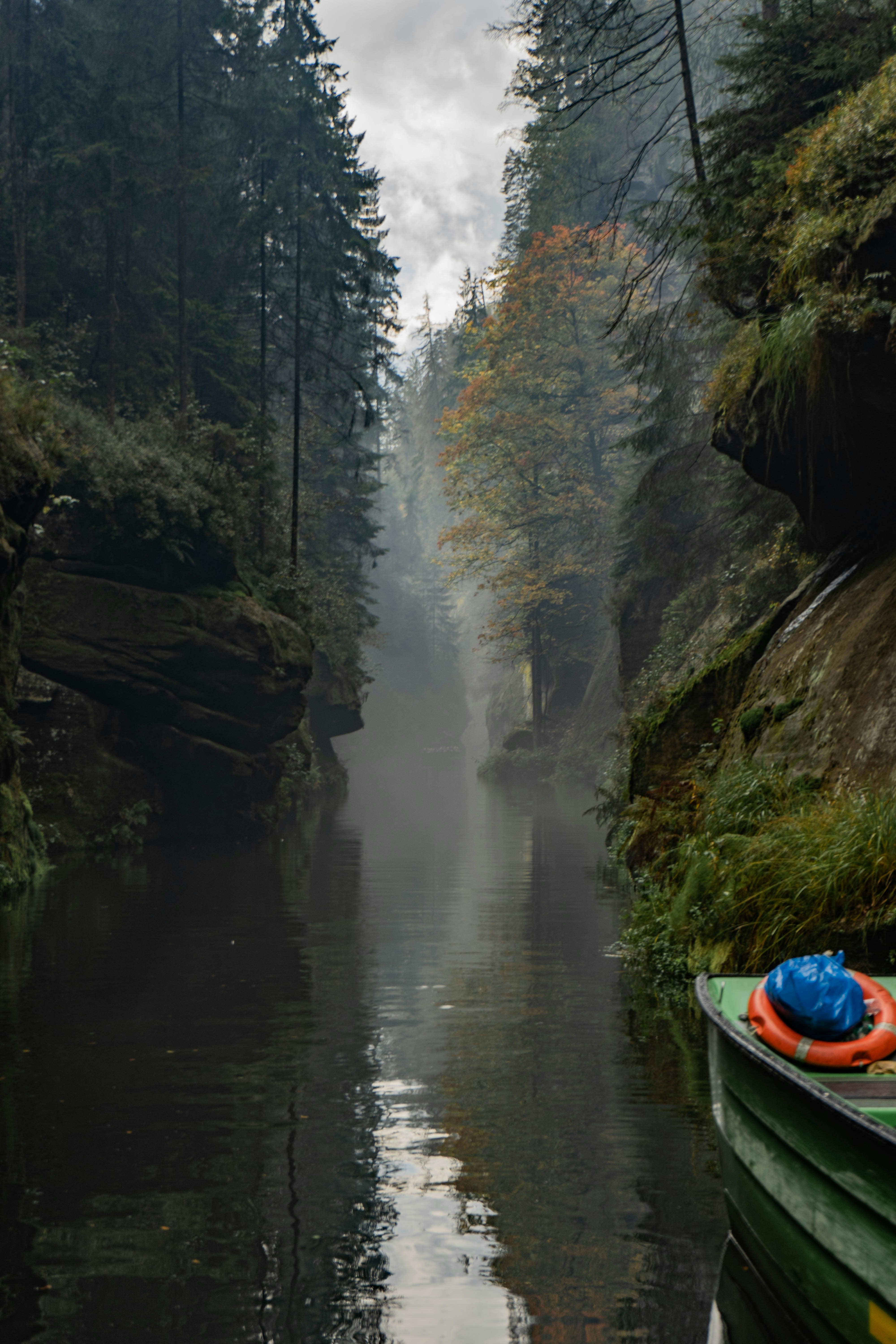 Serene waterway flanked by towering cliffs and autumn foliage, shrouded in mist. A boat rests quietly at the edge, inviting exploration.