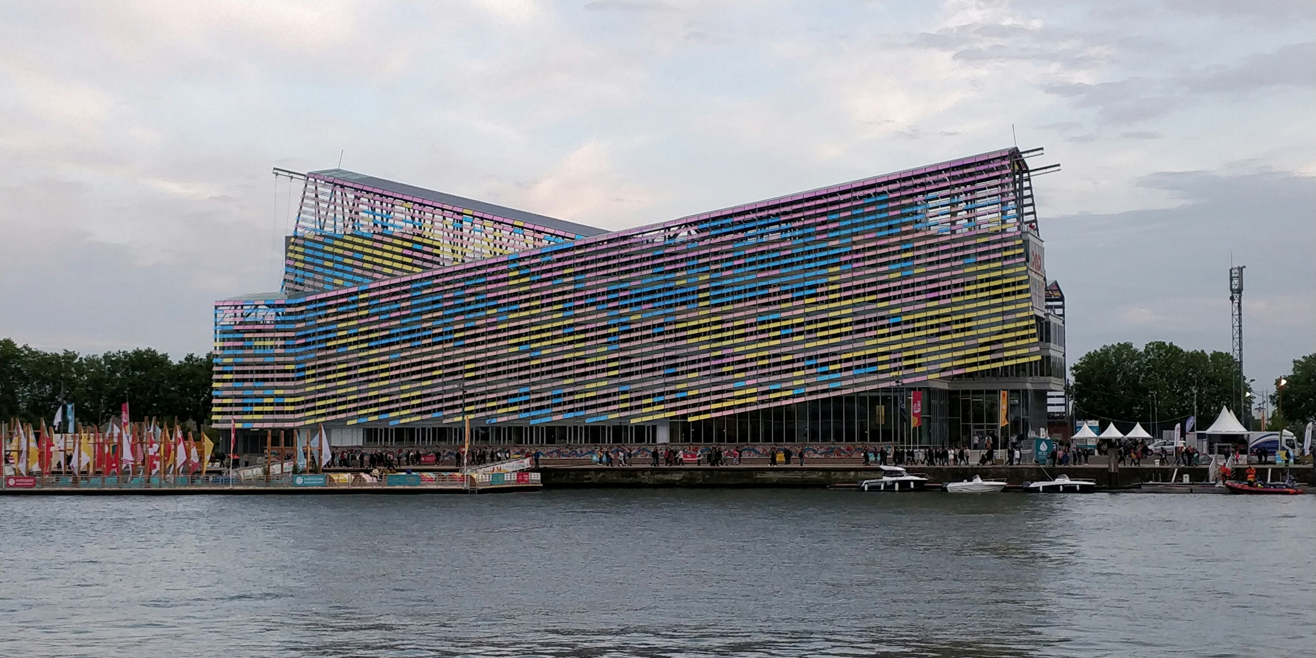 Vibrantly patterned building with angular roof beside a calm waterfront under a cloudy sky.