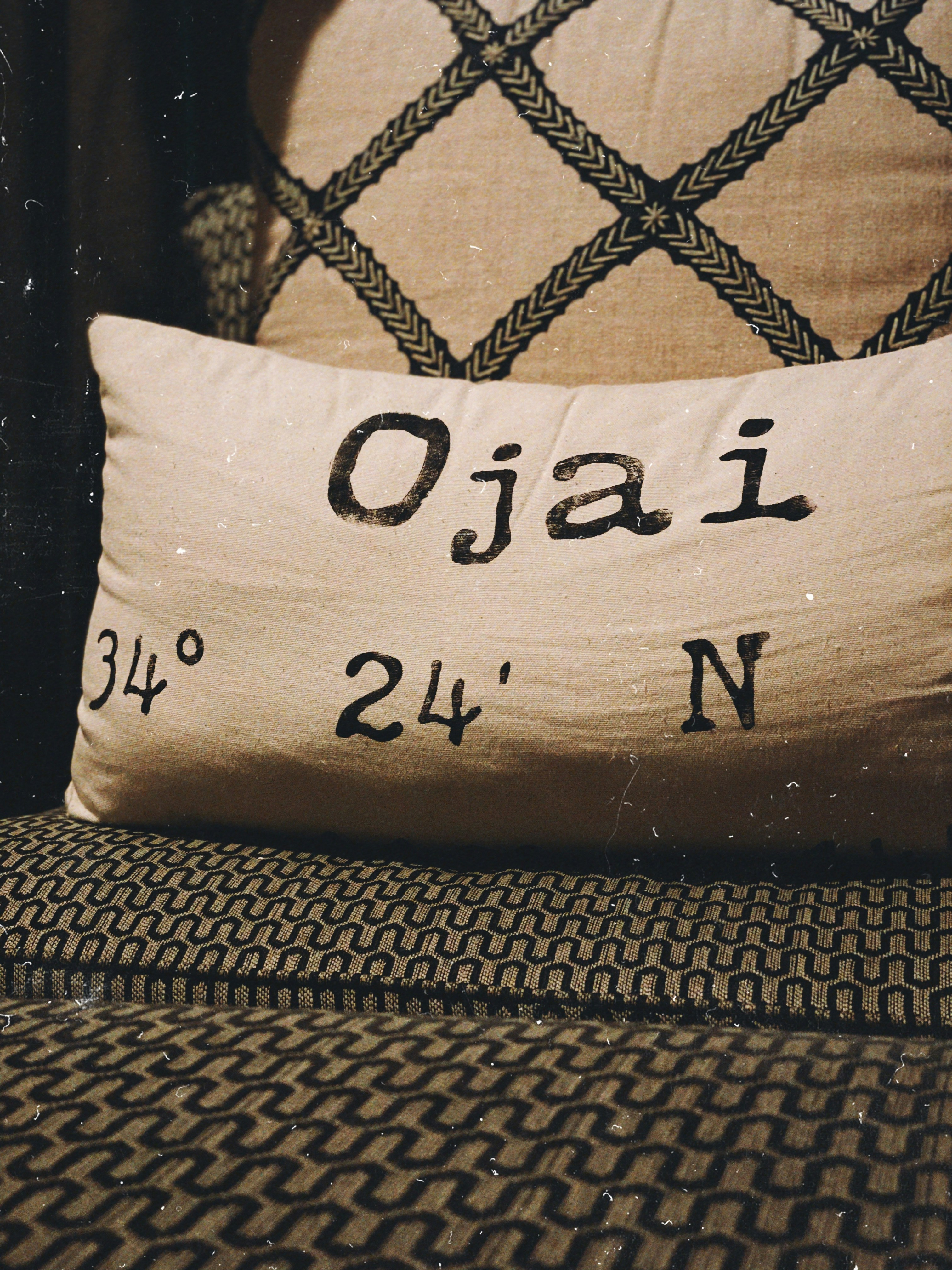 Photograph of a beige throw pillow printed with 'Ojai' and the coordinates 34° 24' N, resting on a textured geometric-weave surface.