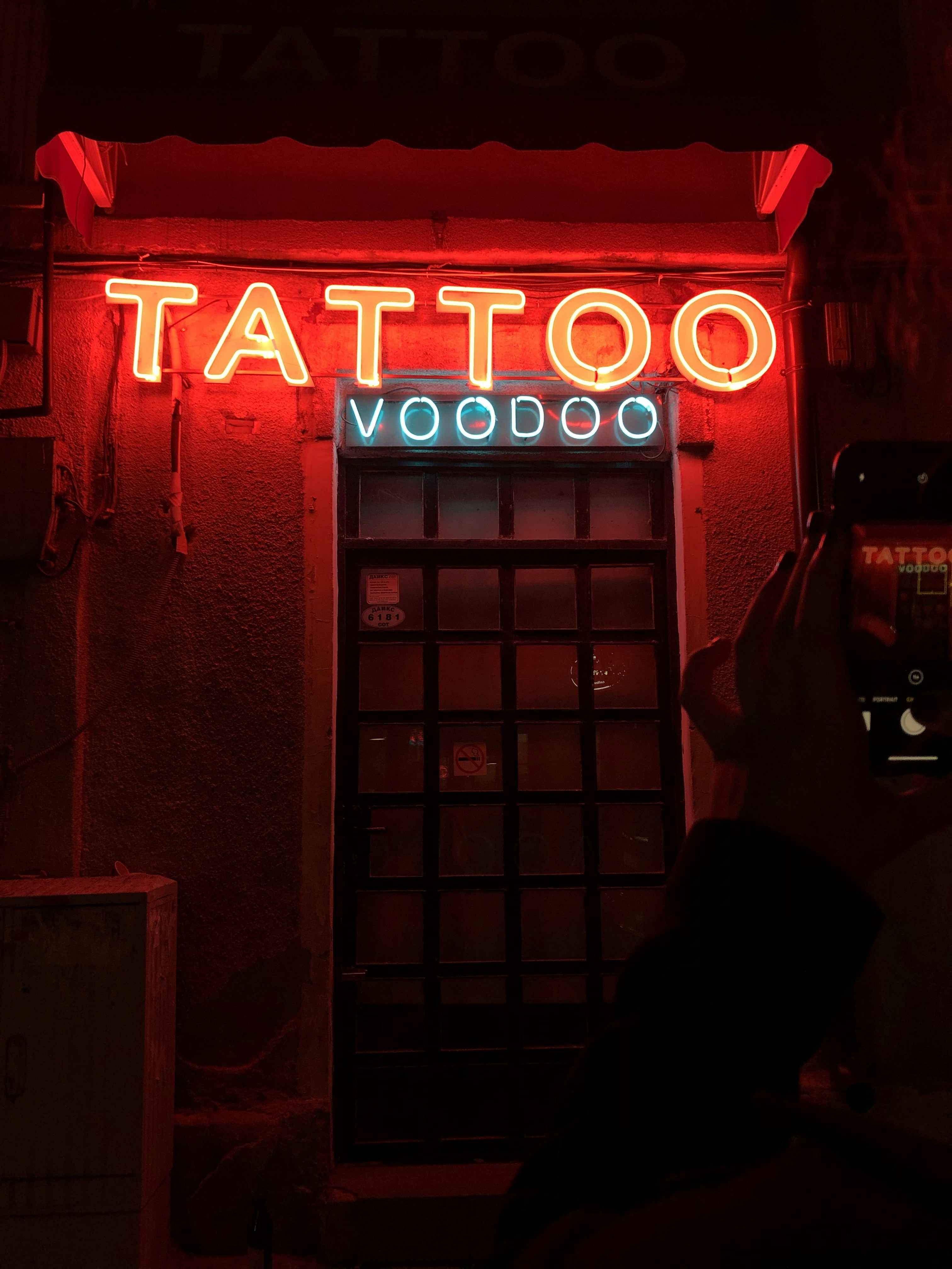 Neon sign reading 'TATTOO VOODOO' illuminates a storefront at night, contrasting with the dark surroundings.