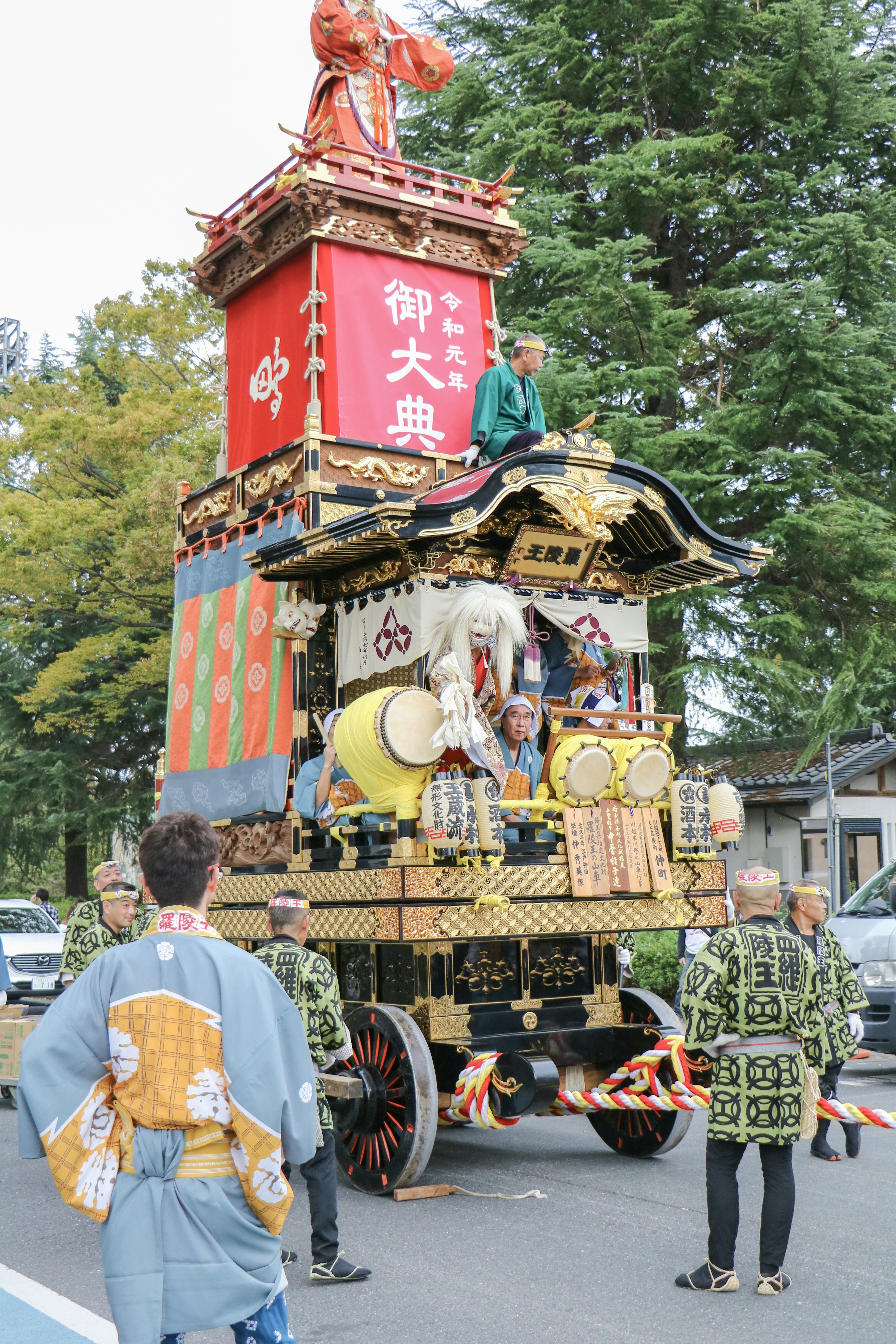 Photograph of an ornate Japanese festival float with drummers, a carved canopy, and a red banner, surrounded by attendants.