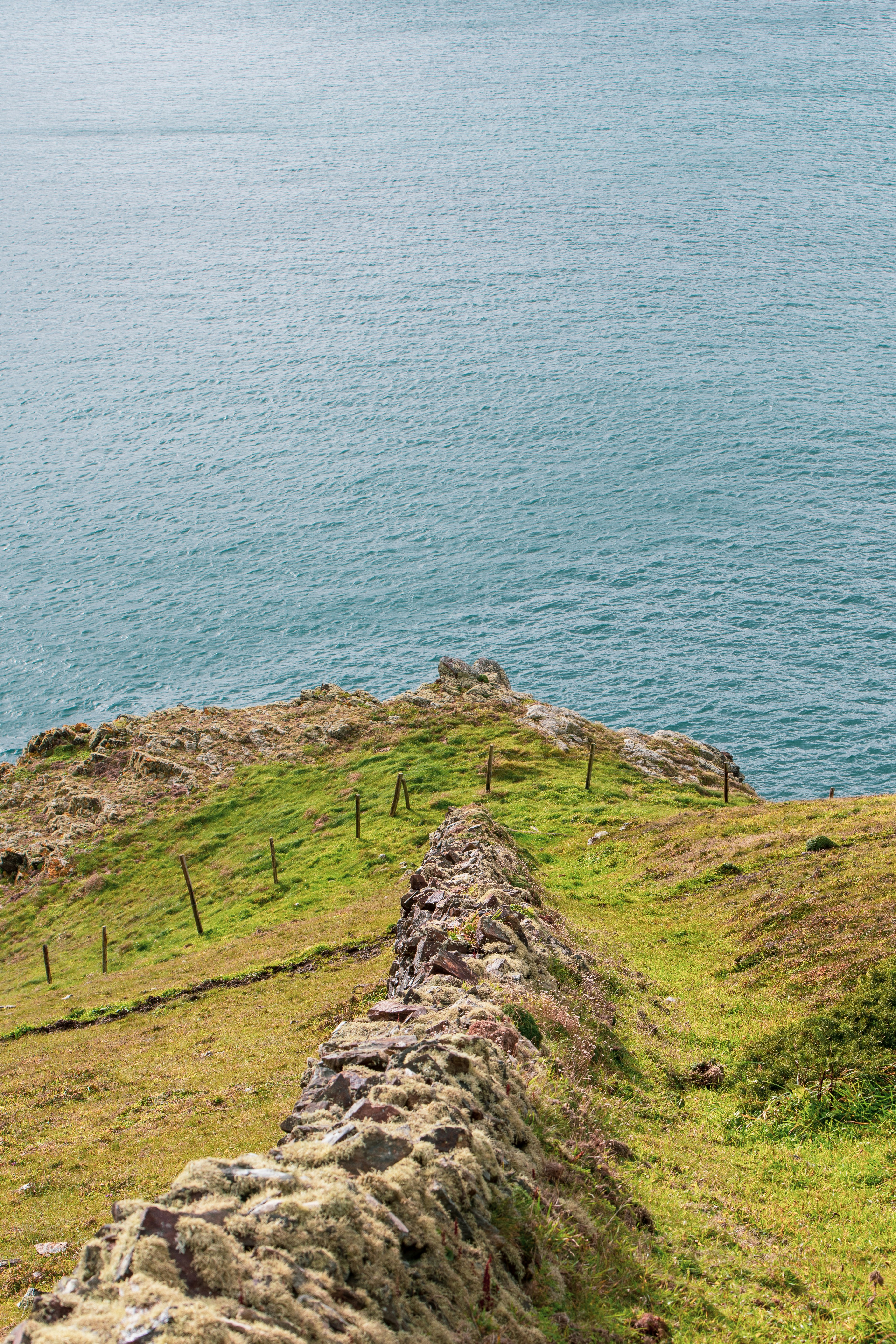 Lush green hillside meets the tranquil blue sea, framed by rustic stone fencing. The scene captures the harmony between land and water.