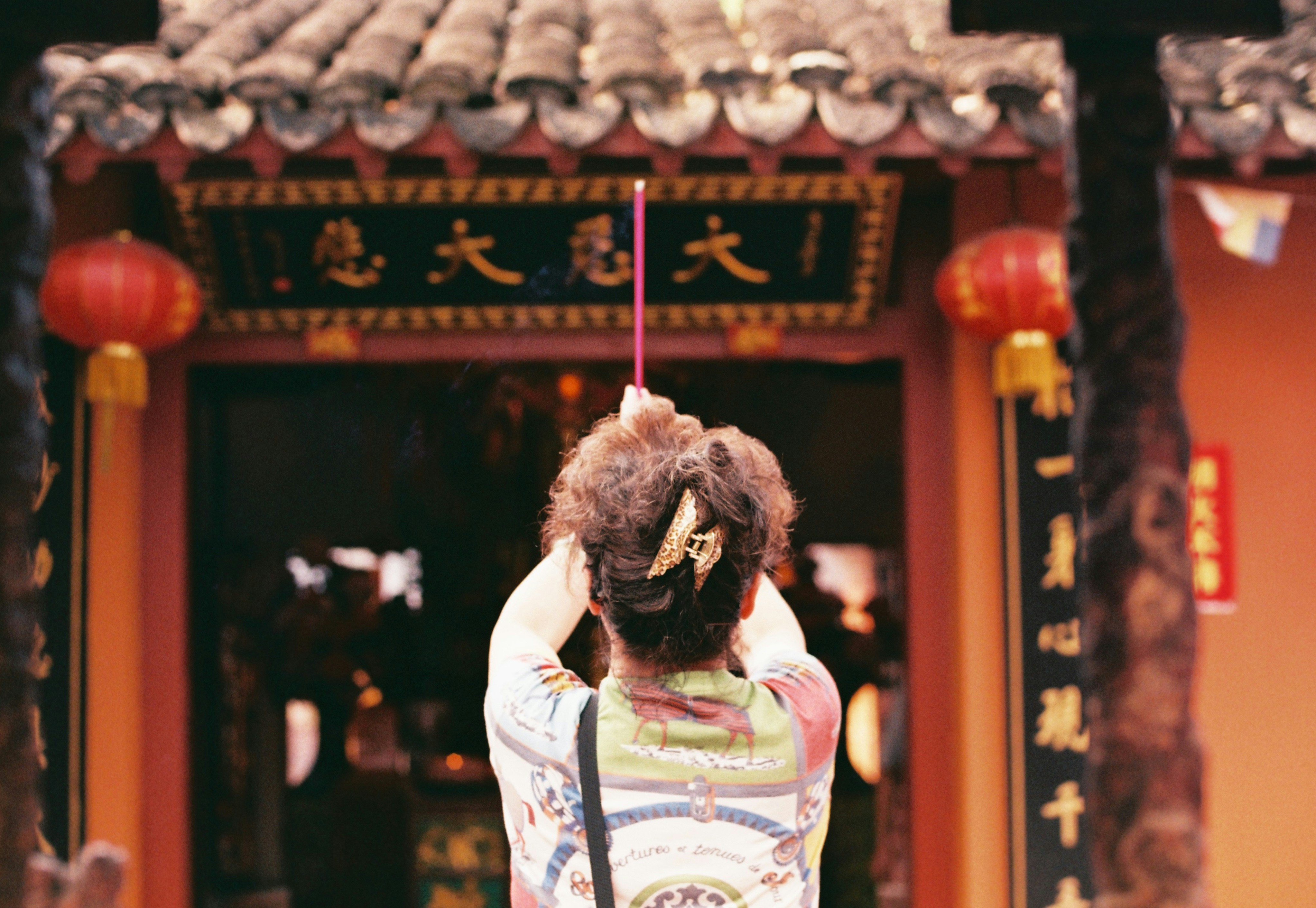 Back-turned traveler in colorful attire stands before a temple doorway, raising a pink straw toward the entrance. Red lanterns frame the ornate signage and tiled roof, creating a ceremonial backdrop.
