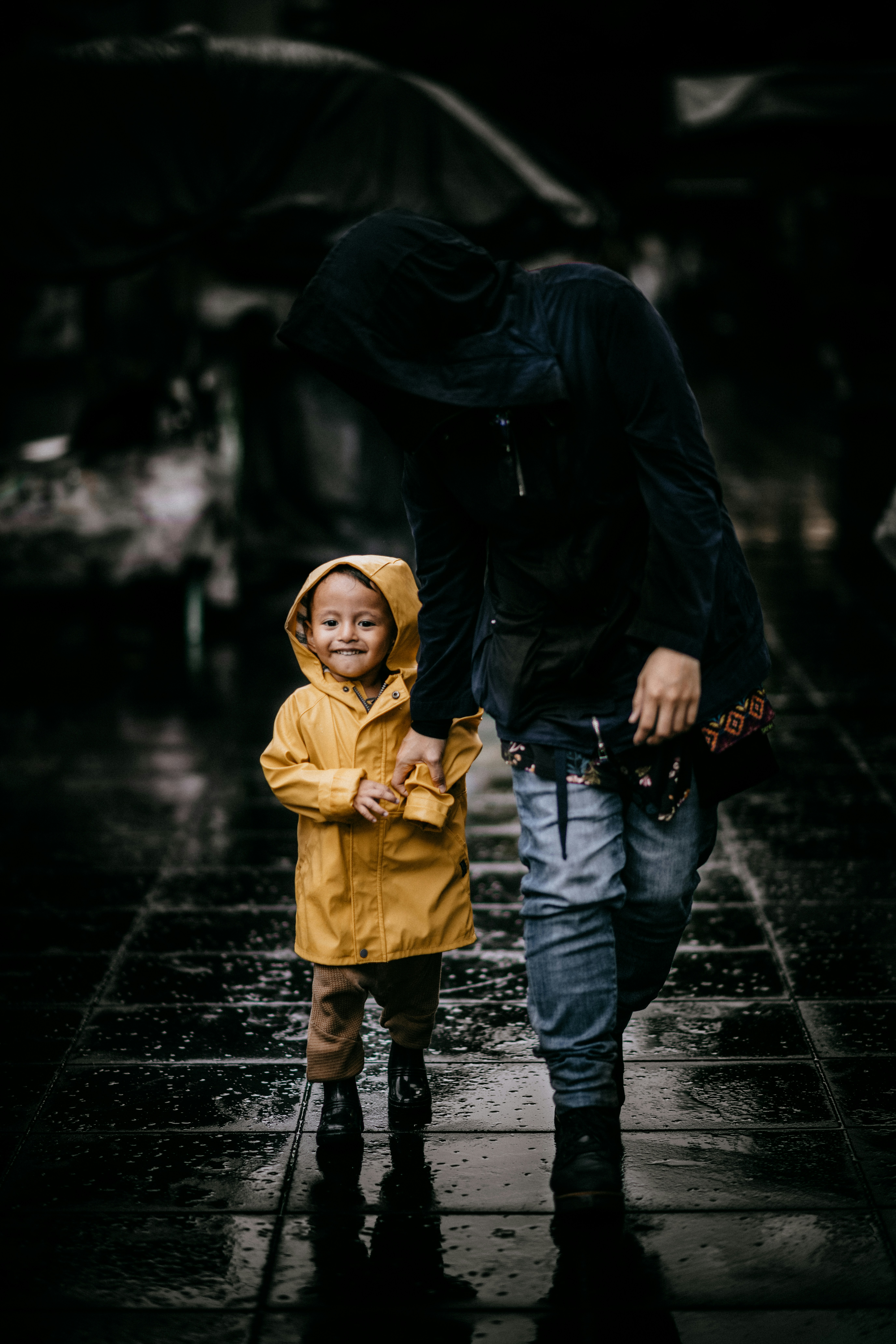 Child in a yellow raincoat holds hands with an adult in a black jacket on a rainy street.