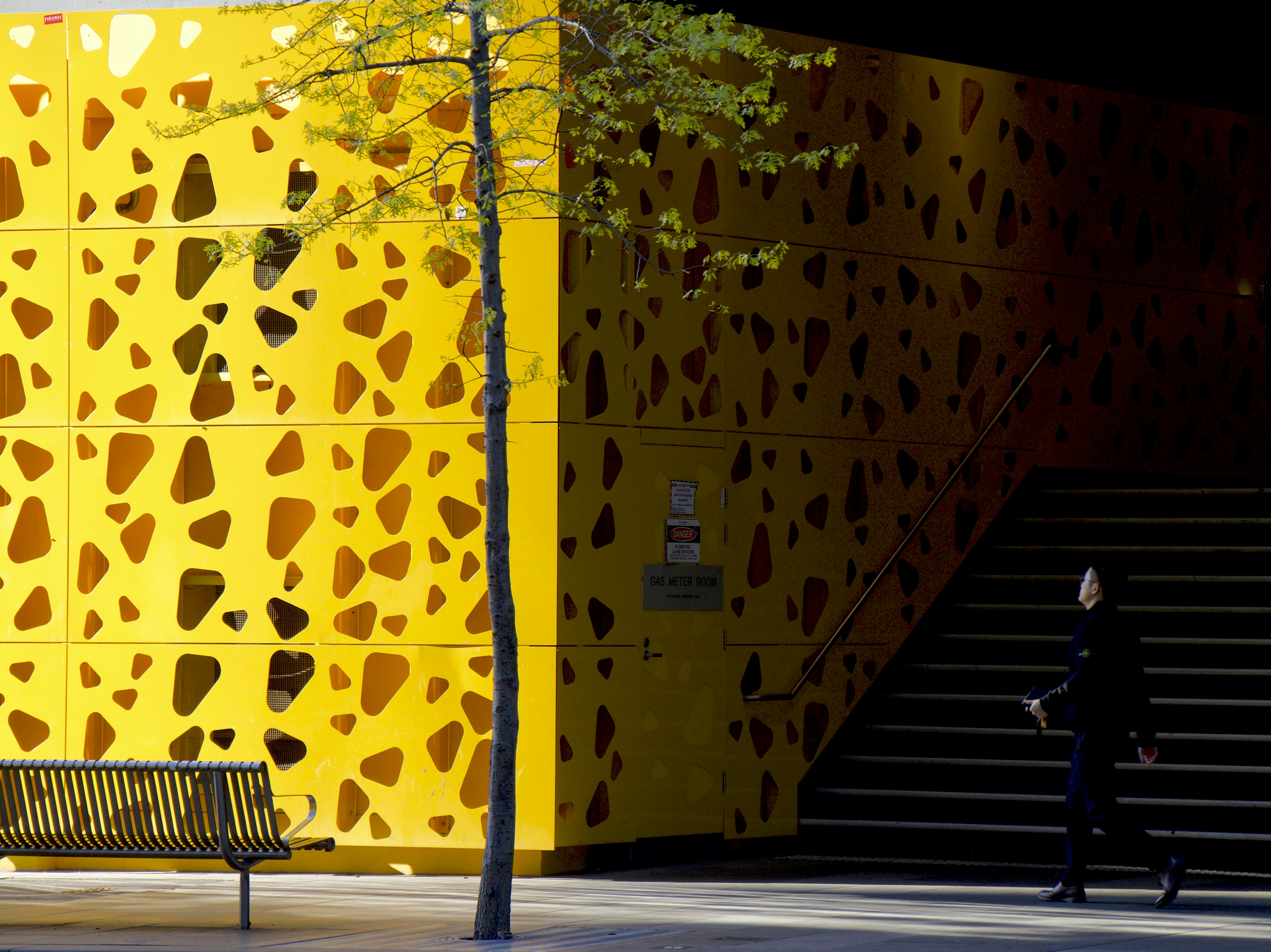 Yellow perforated wall, lone tree, and distant pedestrian at the stairwell