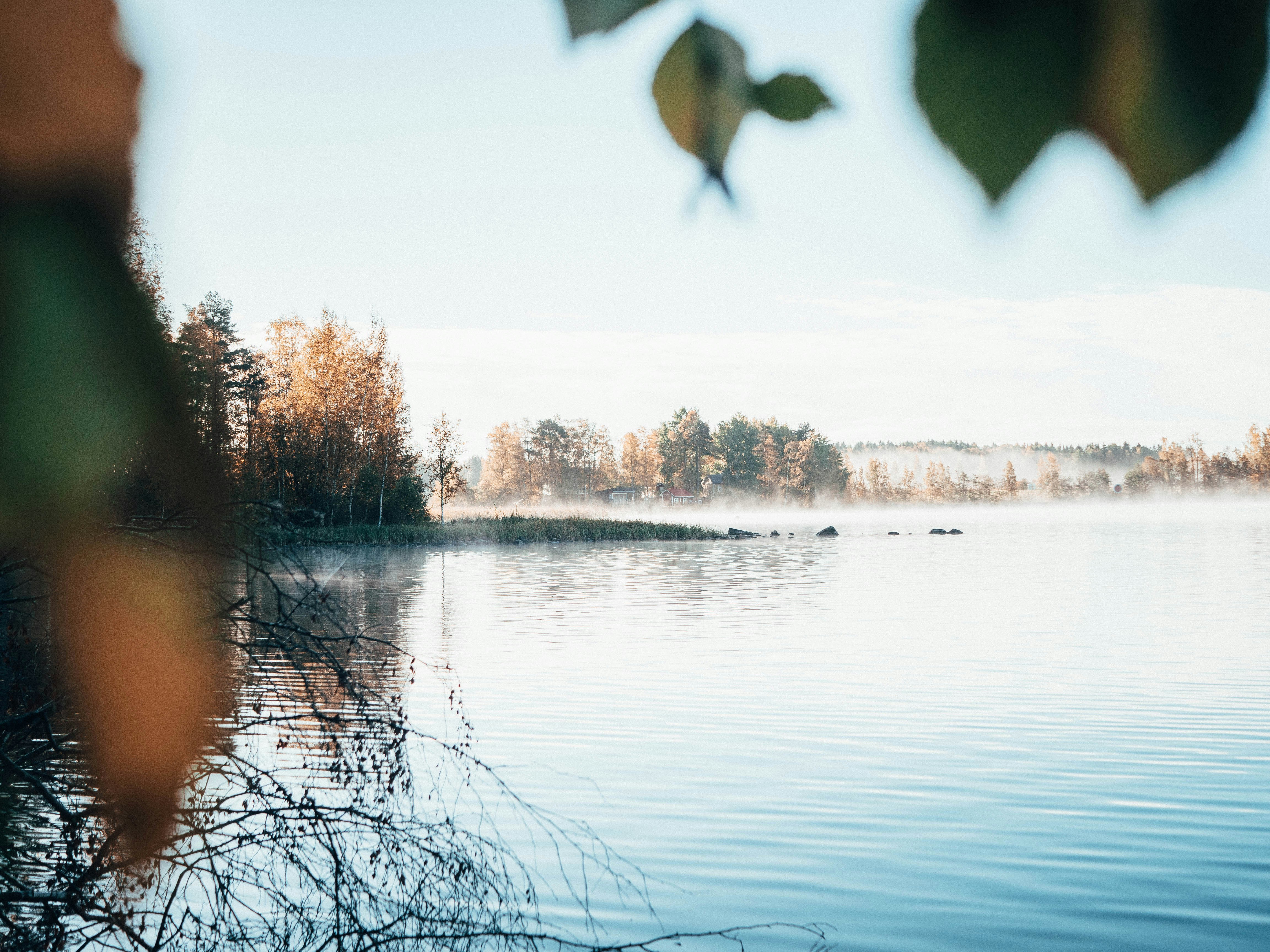 Calm lake reflecting autumn foliage under a clear sky, framed by leaves in the foreground. Mist gently rises from the water's surface.