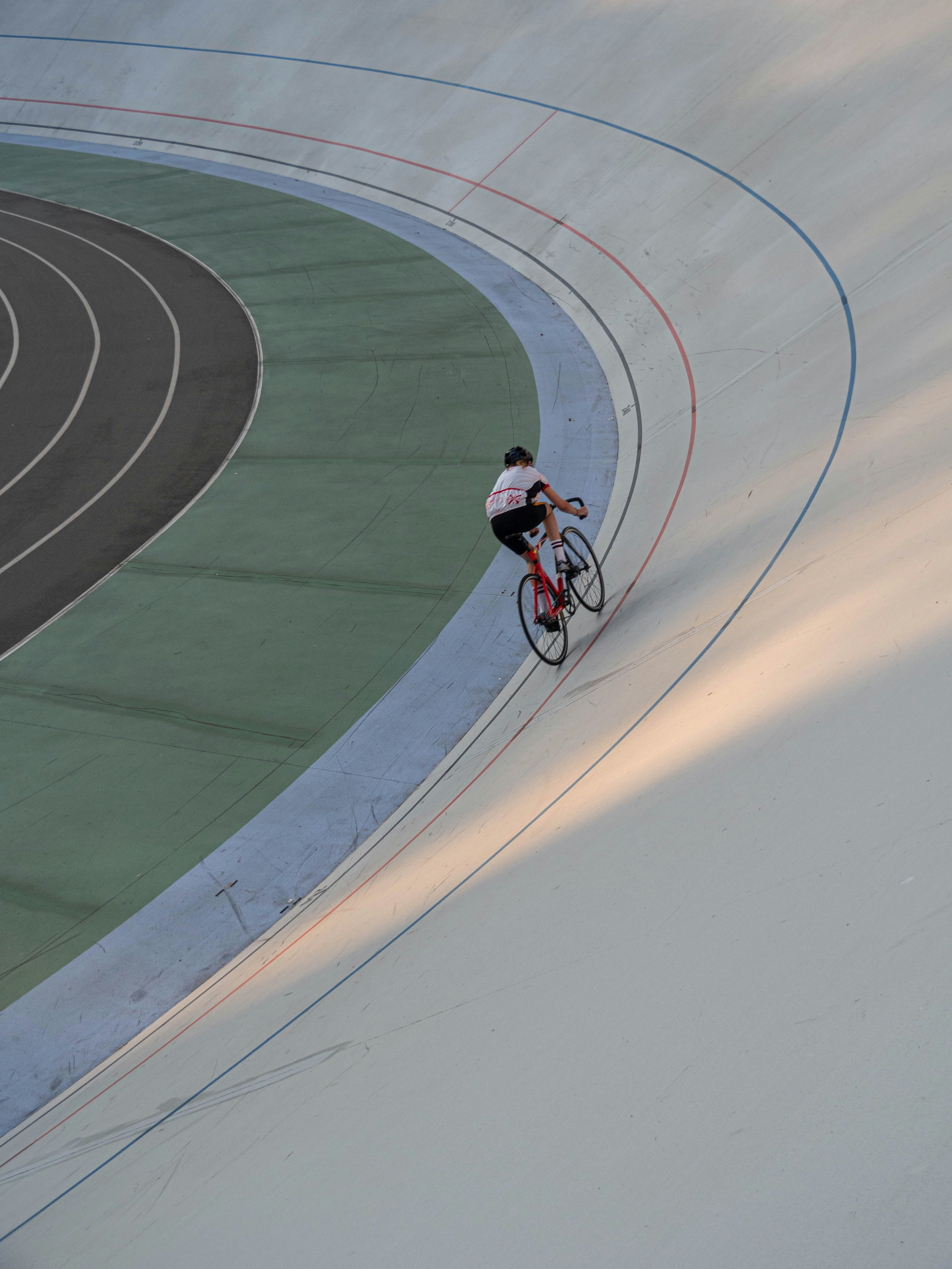Photograph of a cyclist riding up the curved banking of a velodrome, with concentric colored lanes looping around him. The shot emphasizes geometry and motion.