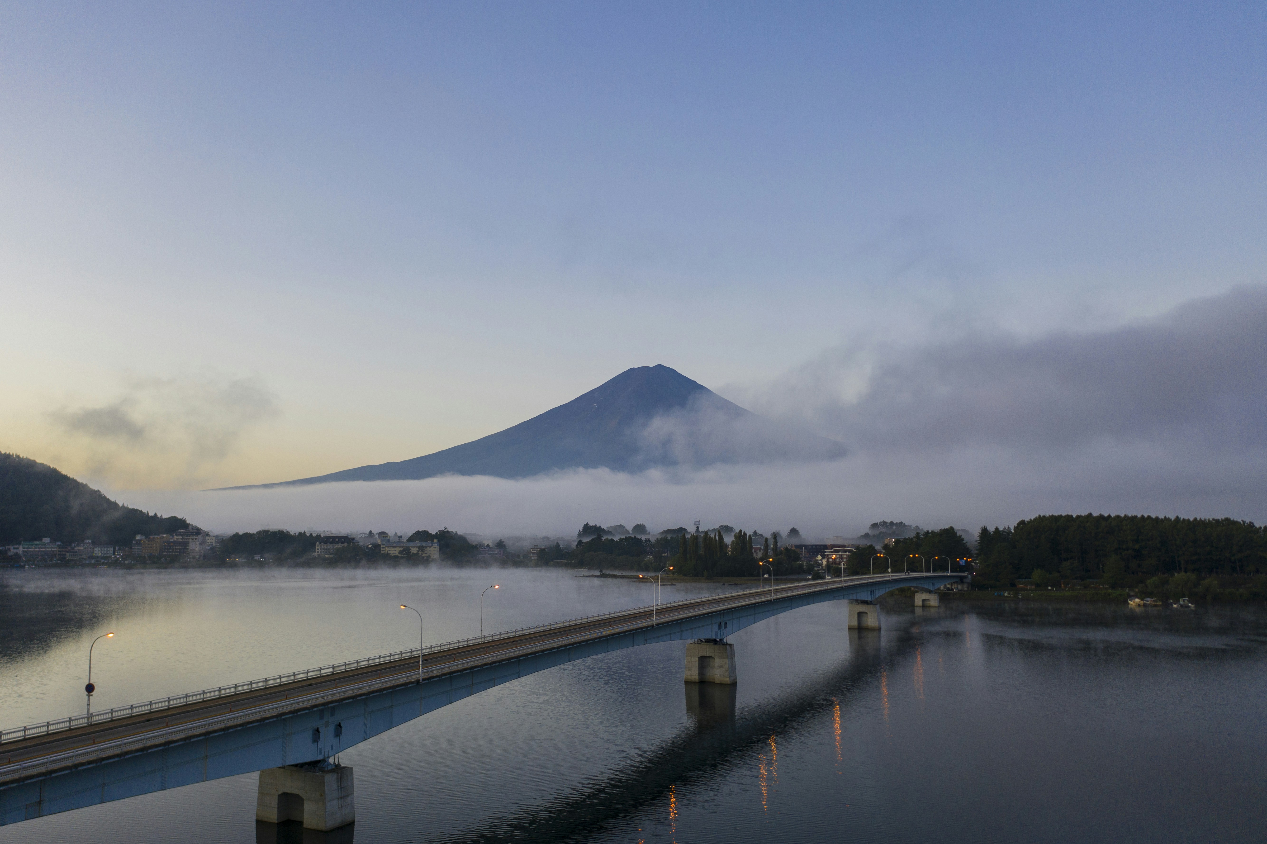 Mount Fuji rising above a tranquil lake with a bridge in the foreground under a clear sky.