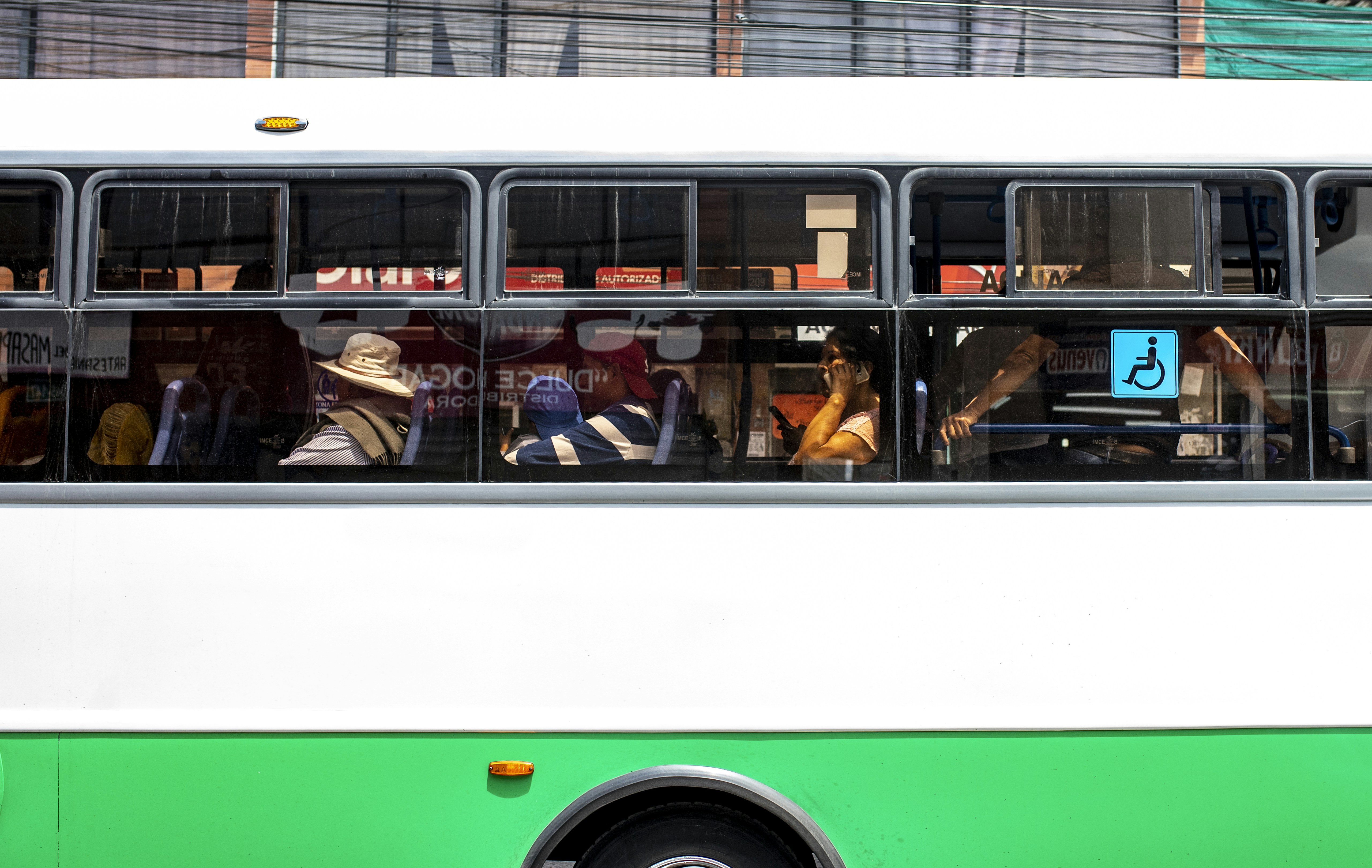 Passengers seated inside a city bus, showcasing a mix of individuals and their unique expressions. The bus features a green and white design with a visible accessibility symbol.