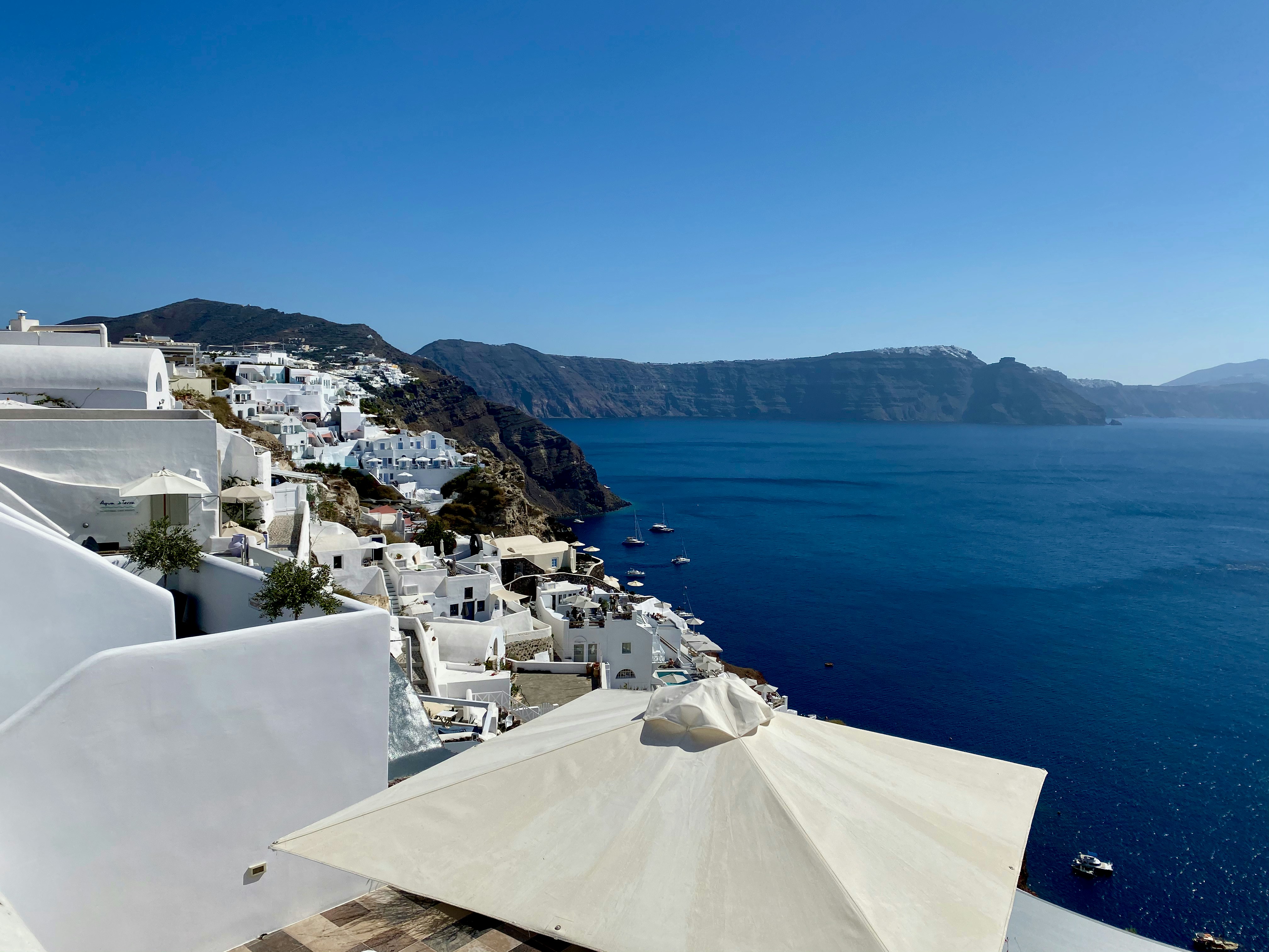 Whitewashed buildings cascade down cliffs overlooking the deep blue Aegean Sea, with a sunshade in the foreground adding to the tranquil atmosphere.