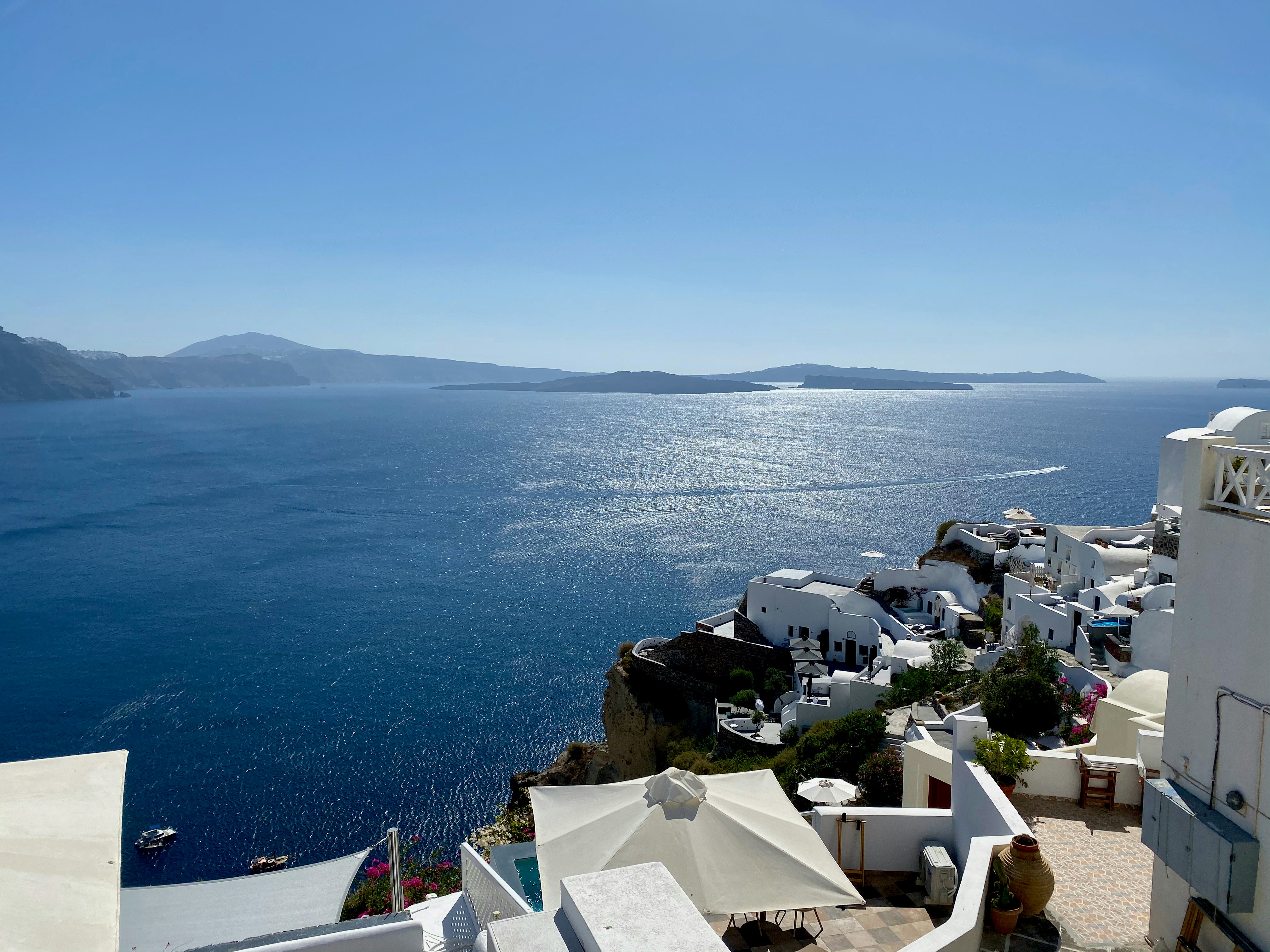 Whitewashed buildings cascade down a cliffside overlooking the shimmering blue sea, with distant islands visible on the horizon.