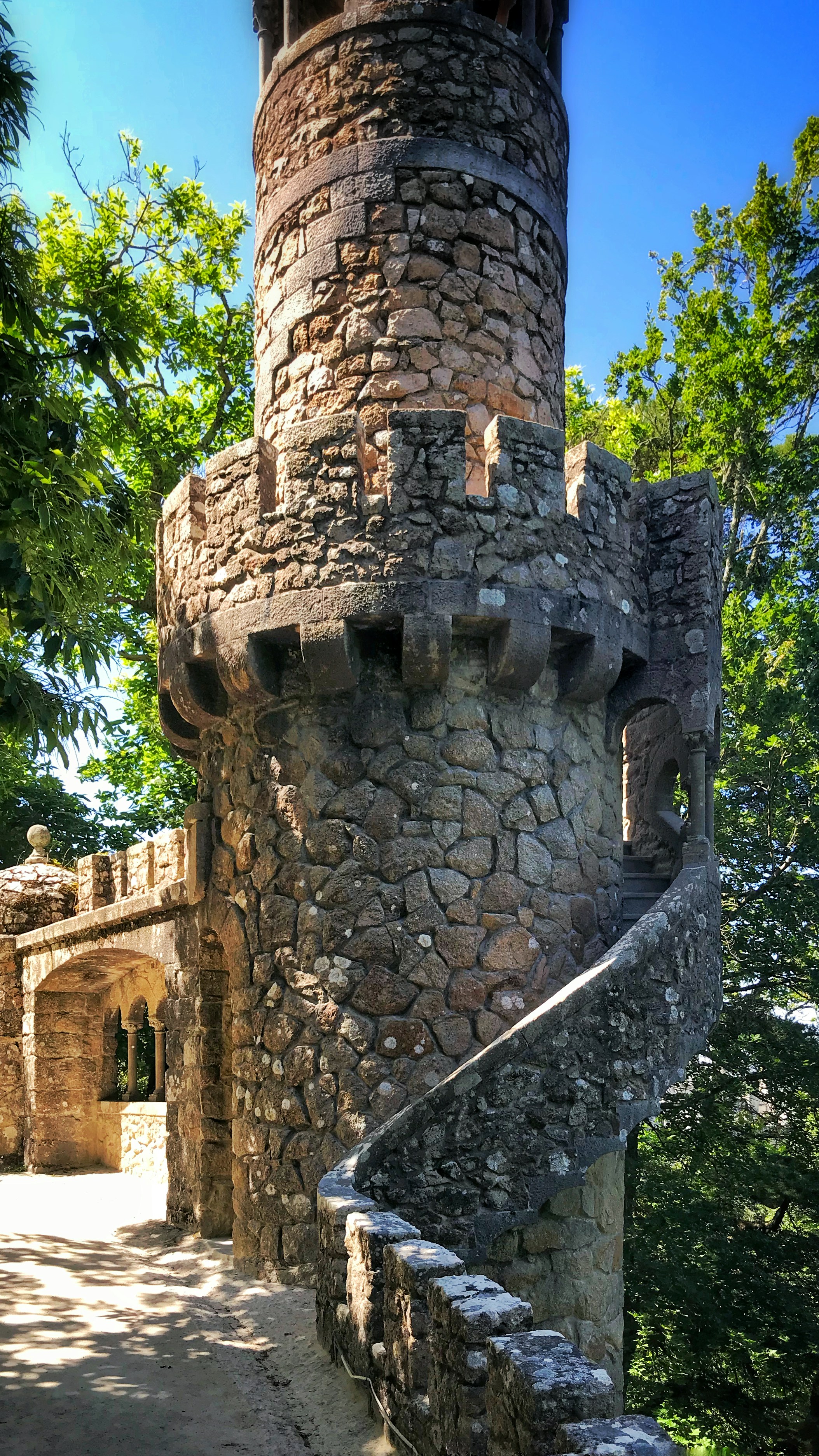 A historic stone tower with a spiraling staircase, surrounded by lush greenery and blue skies. The structure showcases intricate stonework and architectural details.