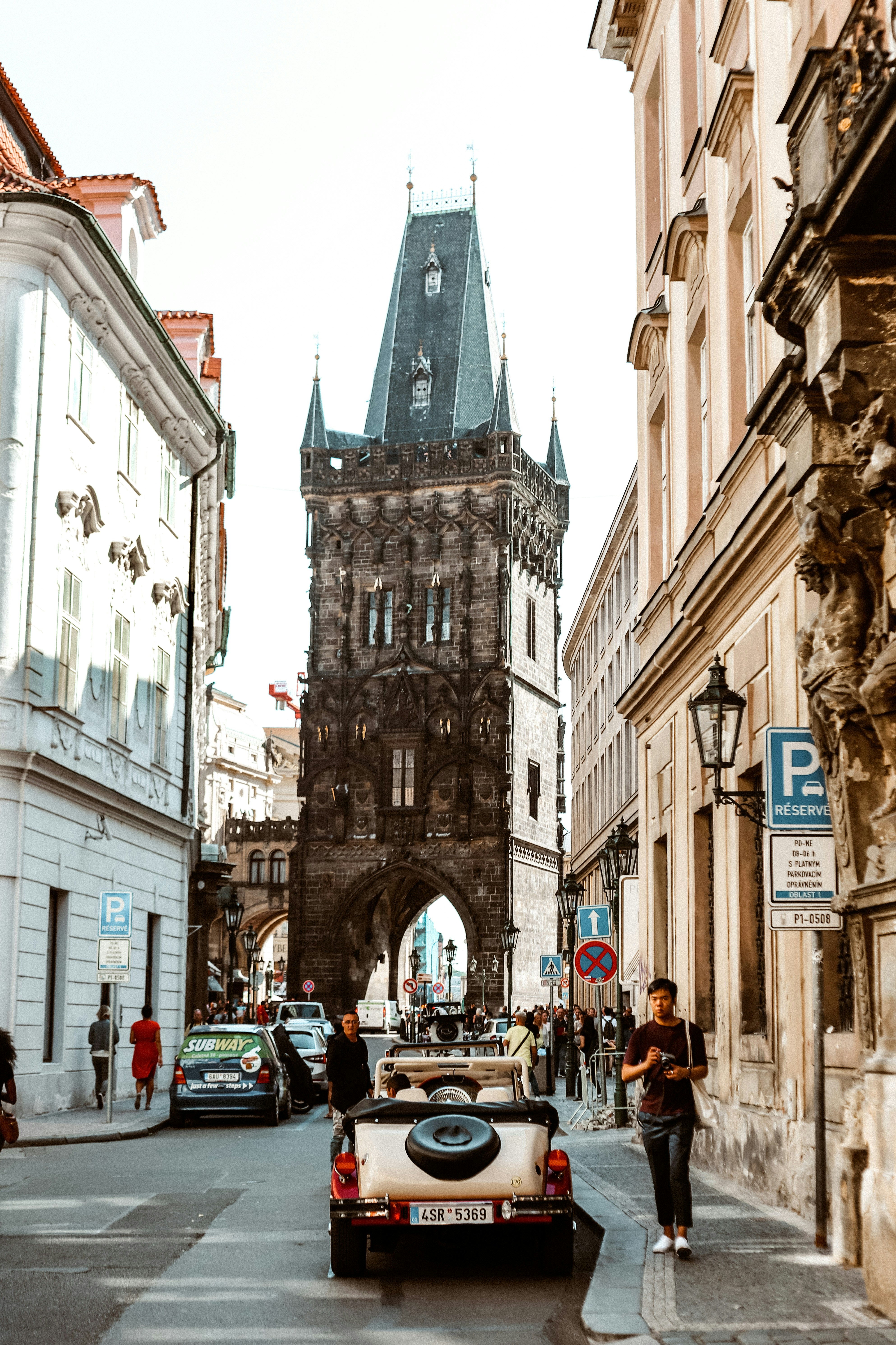 Gothic architecture of a historic tower framed by bustling street activity, showcasing a blend of modern and classic elements. A vintage car adds charm to the scene.