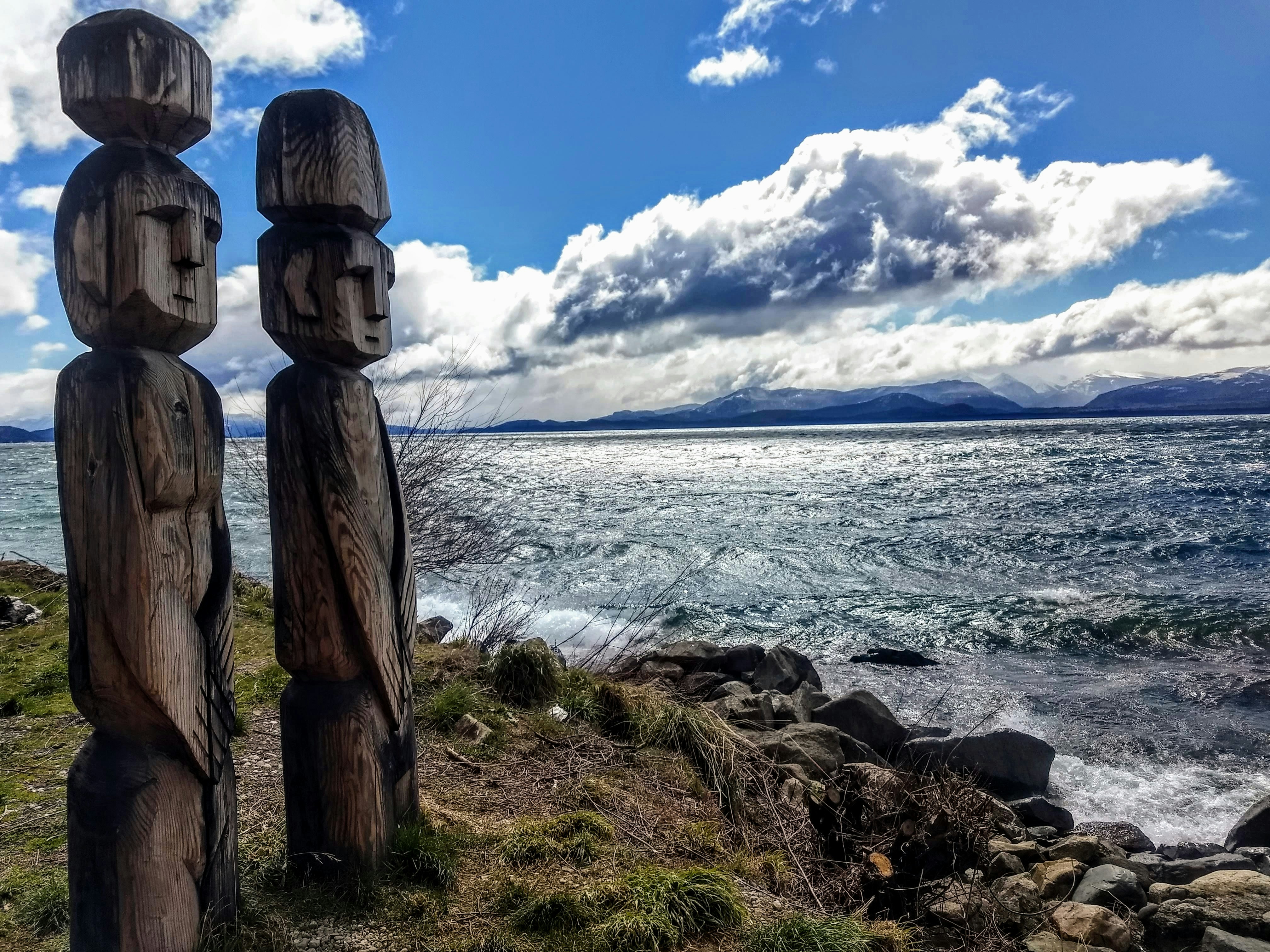 Two intricately carved wooden figures stand watch over a shimmering lake, framed by dramatic clouds and distant mountains.