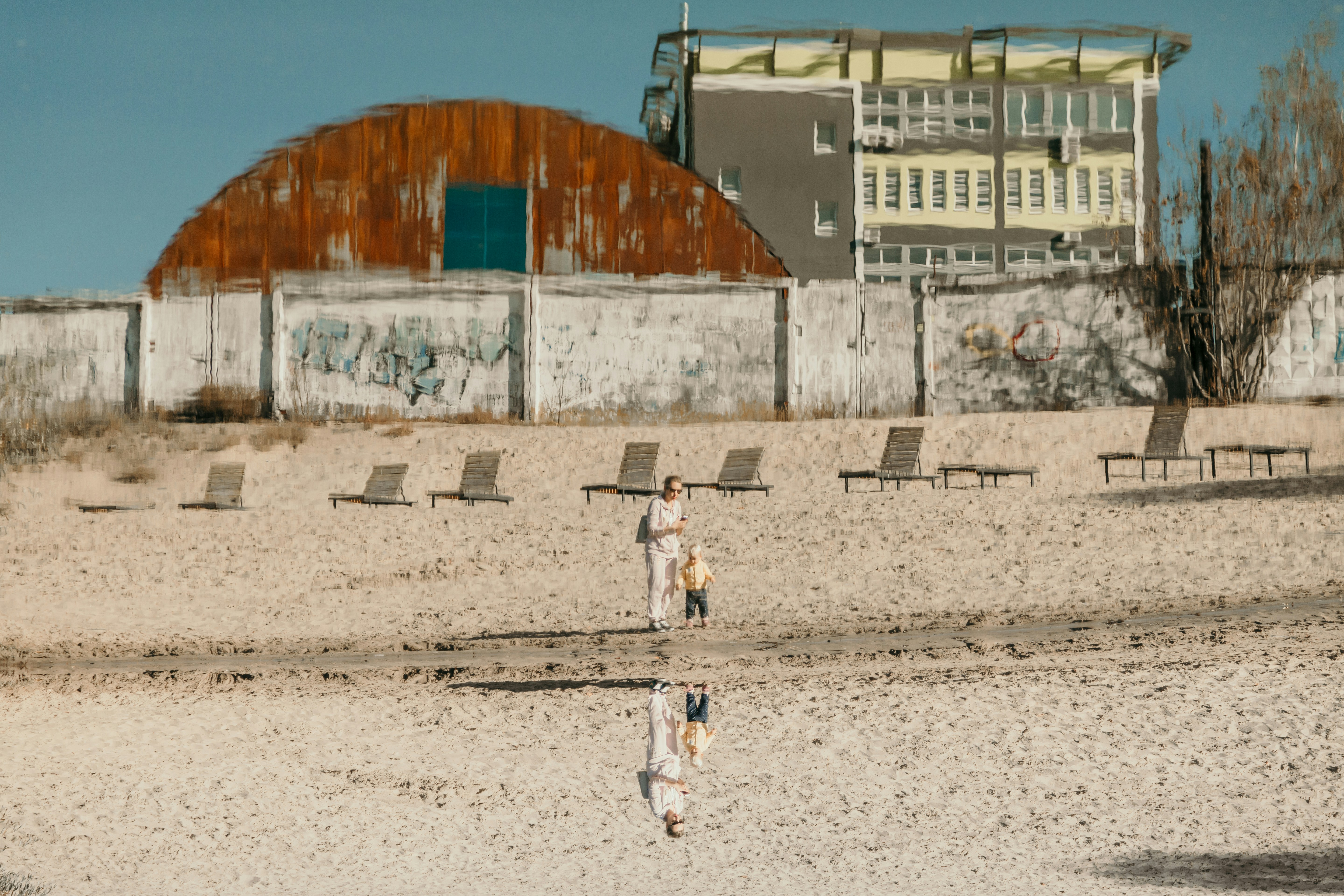 A serene beach scene featuring two figures walking along the shoreline, their reflections mirrored in the calm water. The backdrop includes a distinctive building with a rusted roof.