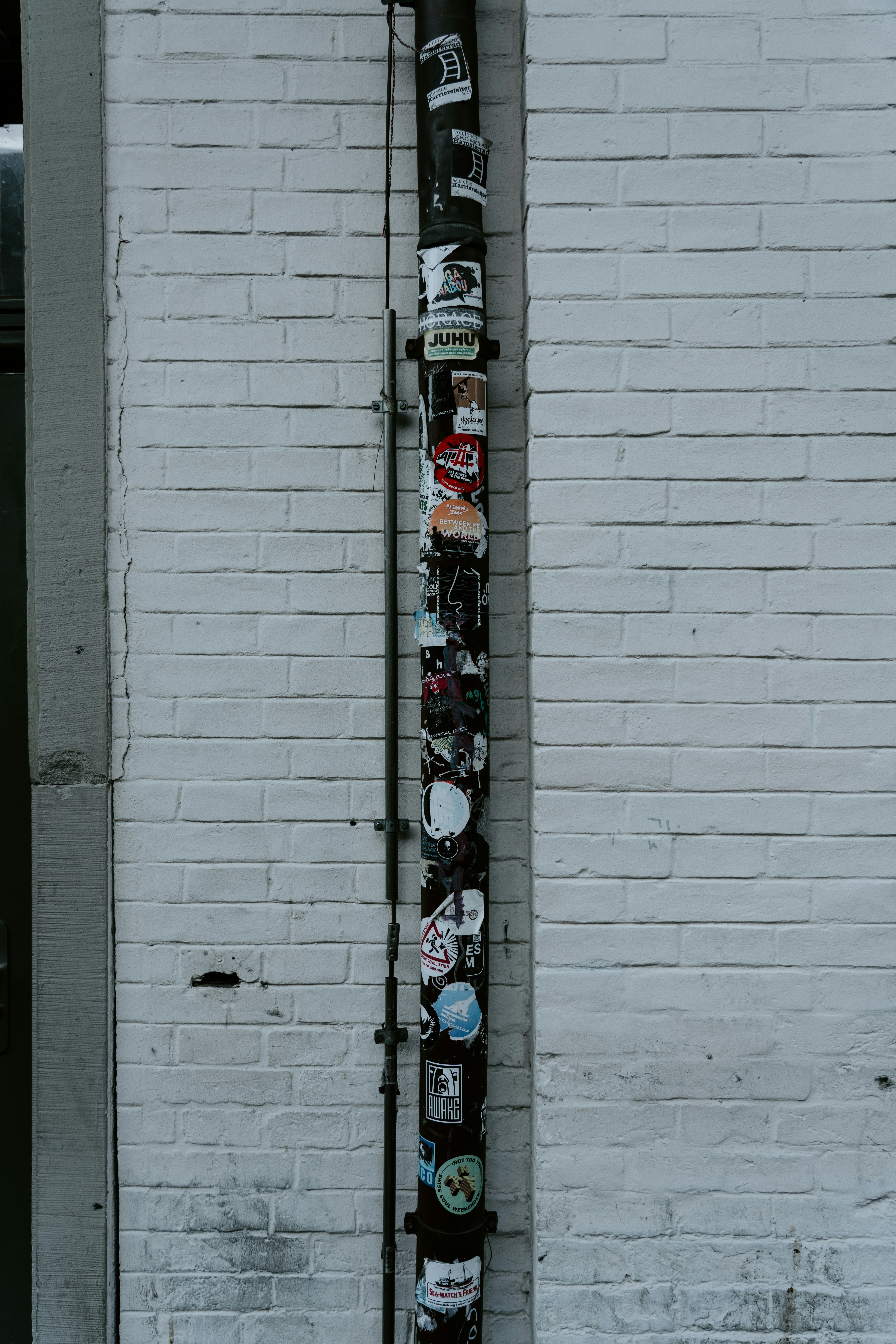 A vertical pipe adorned with a collage of colorful stickers against a white brick wall, highlighting urban art and culture.