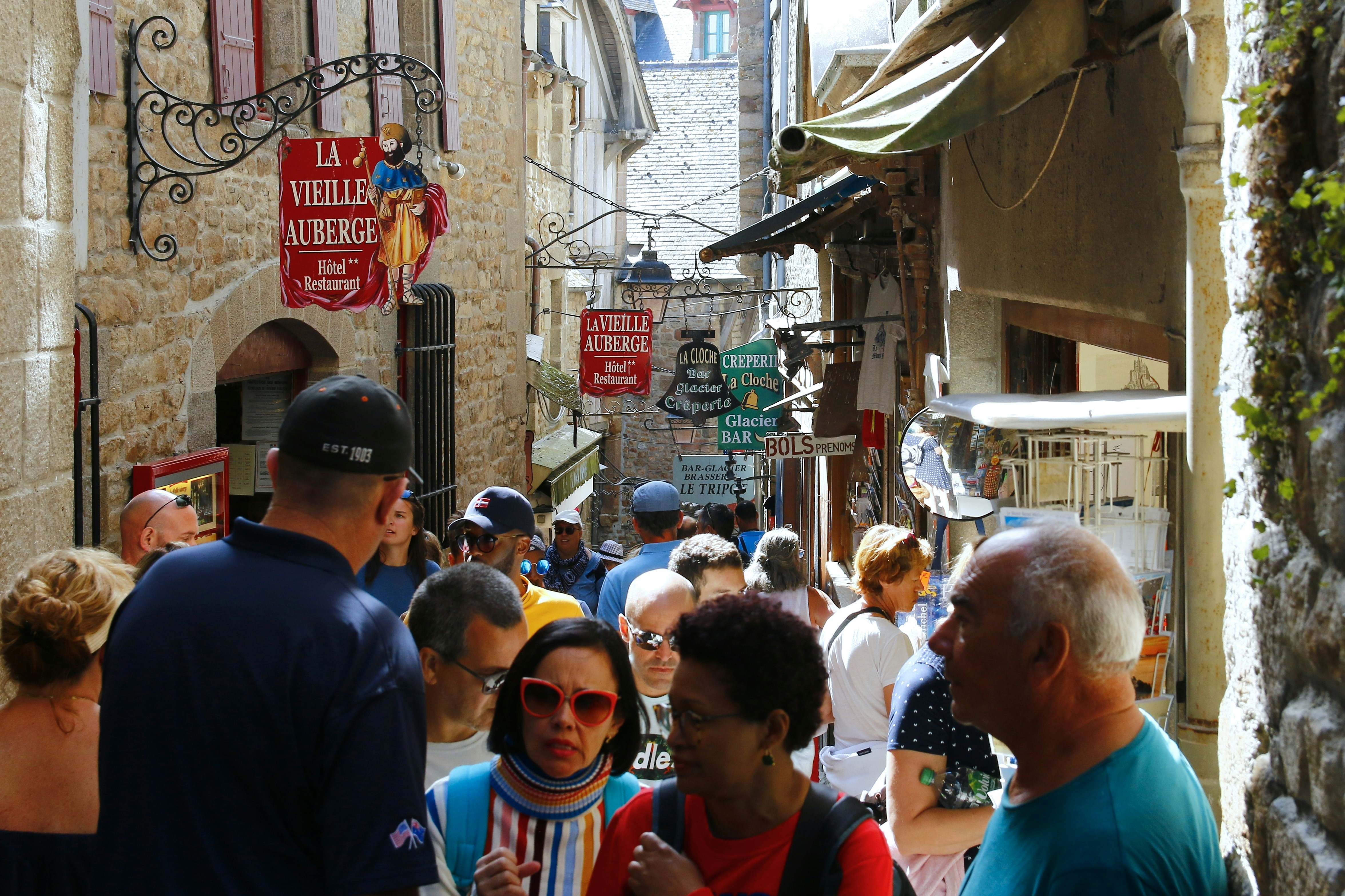 Crowd of tourists navigating a narrow, sunlit alley filled with charming signs and stone walls. The scene captures the essence of local culture and bustling activity.