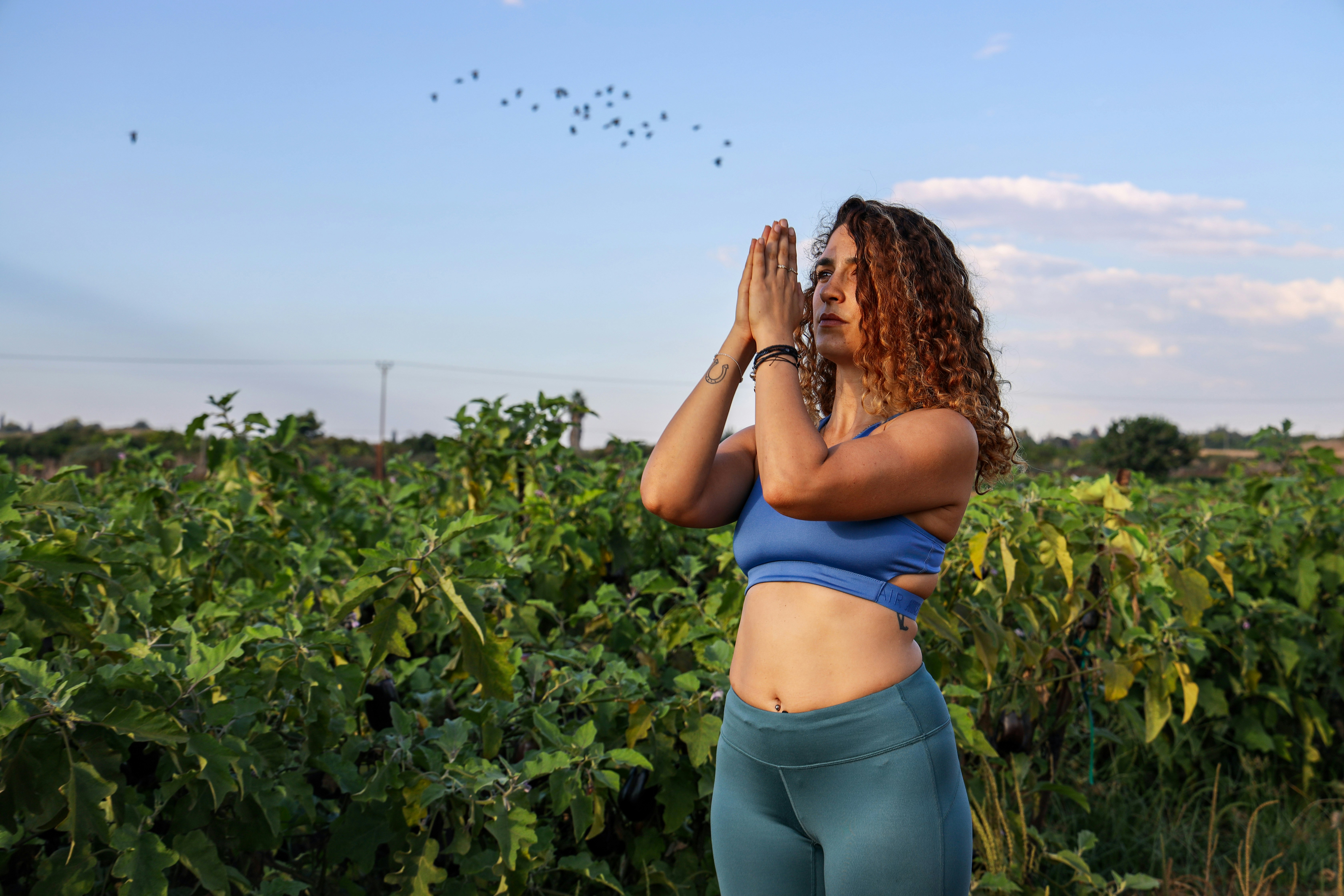 Woman practicing yoga in a lush vineyard under a clear sky, with birds flying in the distance.