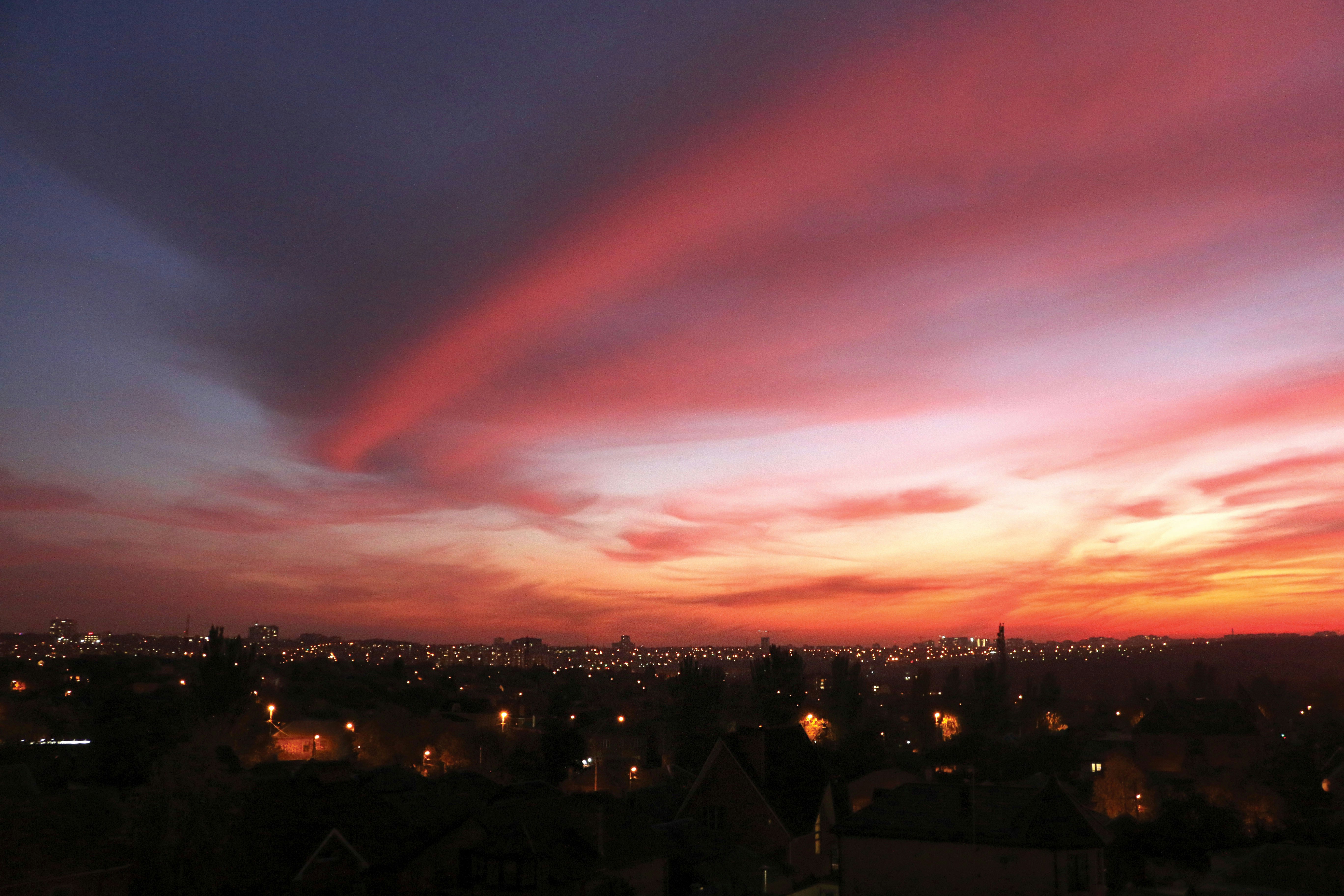 Vibrant sunset hues blend across the sky, illuminating the cityscape below with a warm glow. The scene captures the transition from day to night.