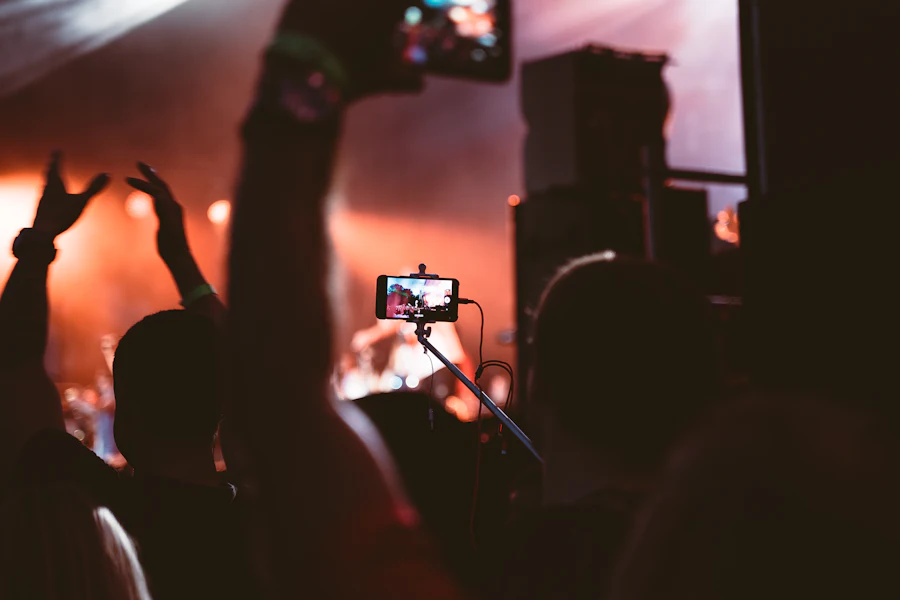 Silhouetted audience members raise their arms while a smartphone on a selfie stick records a brightly lit stage. Warm orange and pink concert lights glow through the haze.