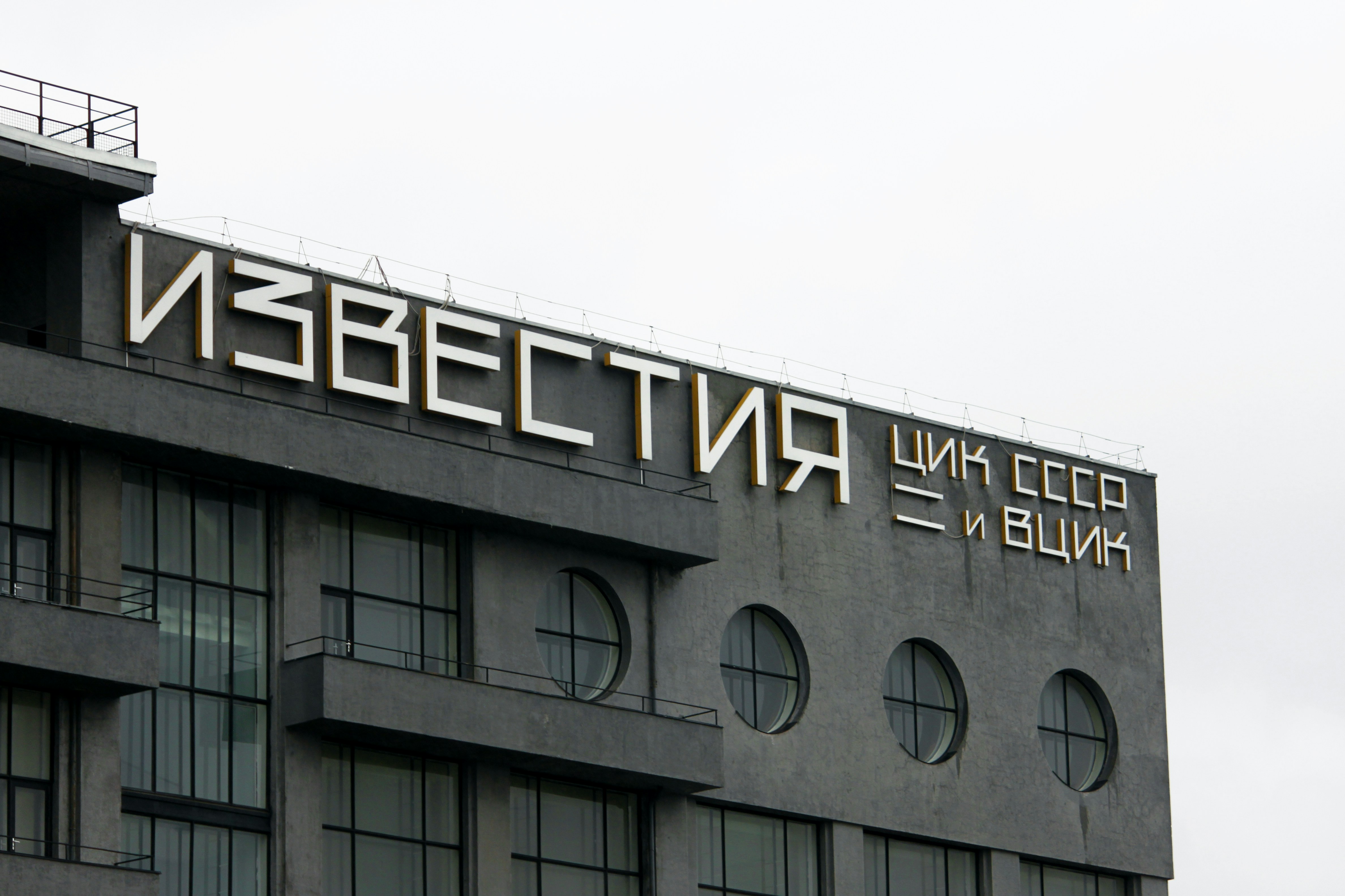 Prominent building facade featuring the word 'Известия' in bold letters, alongside circular windows, showcasing modern architecture against a cloudy sky.