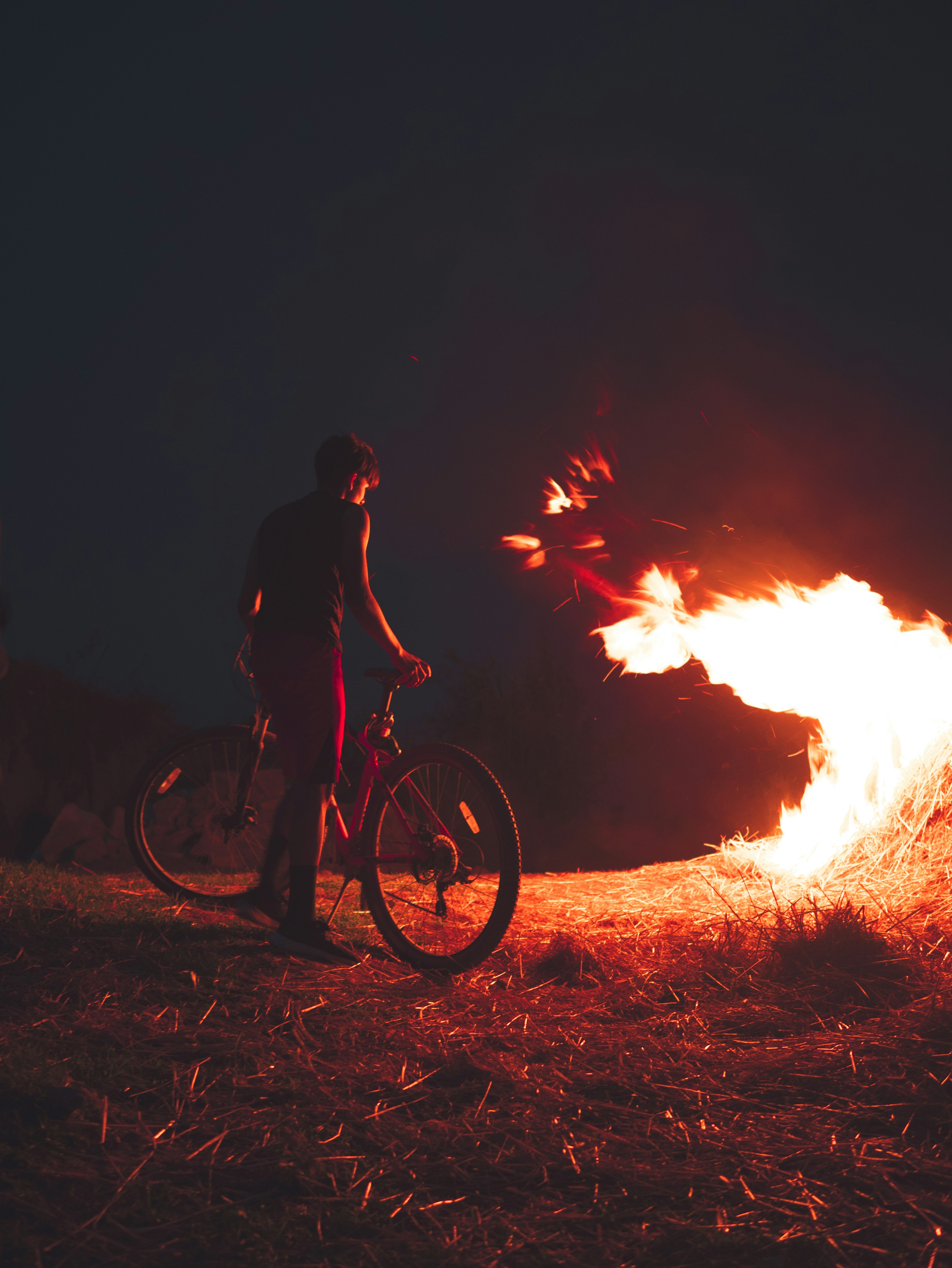 A cyclist stands beside a vibrant fire, creating a striking contrast against the dark night sky. The scene captures the interplay of light and motion.