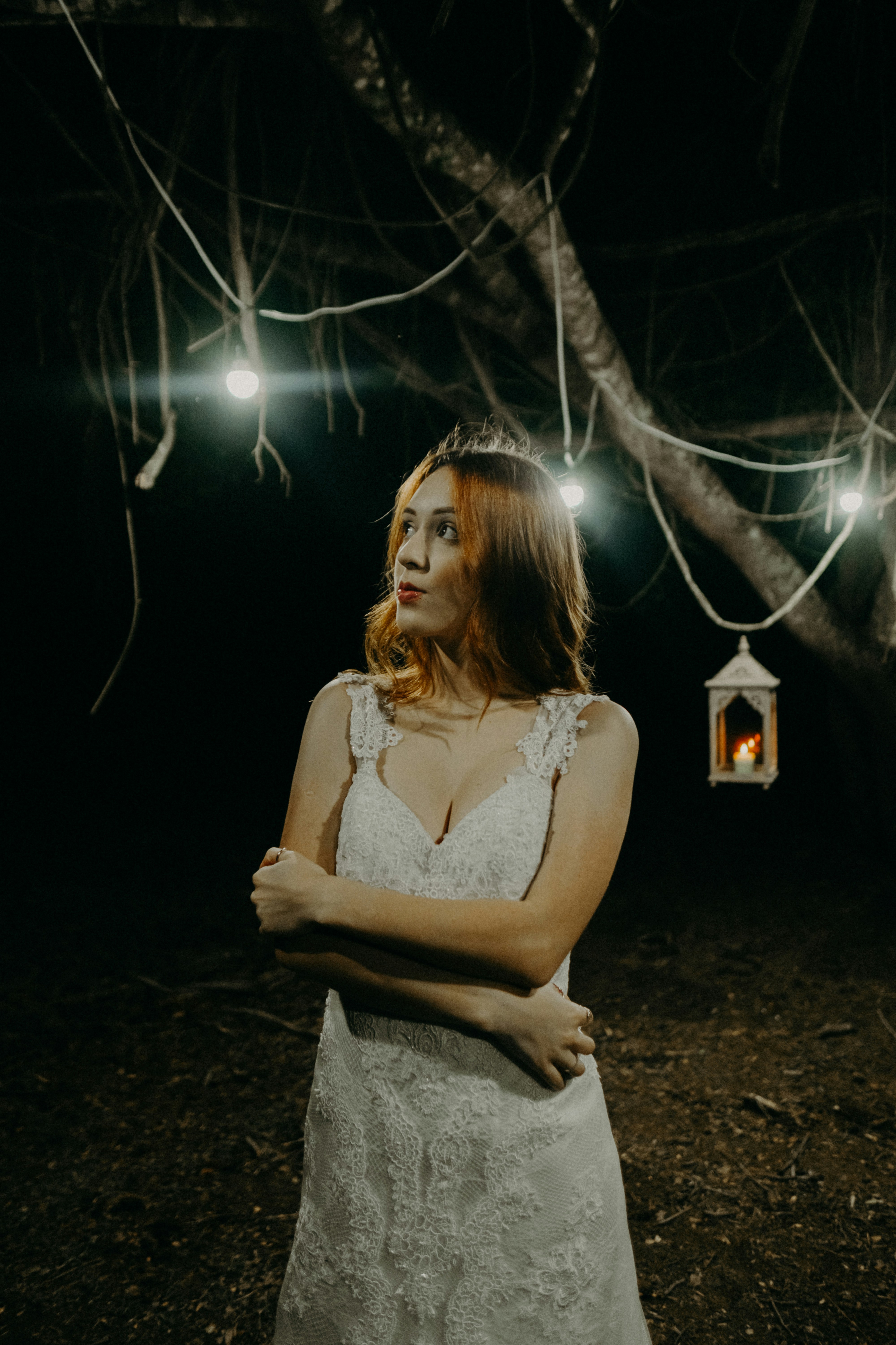 A bride stands under a tree adorned with glowing lanterns, exuding a sense of contemplation and mystery in a darkened setting.