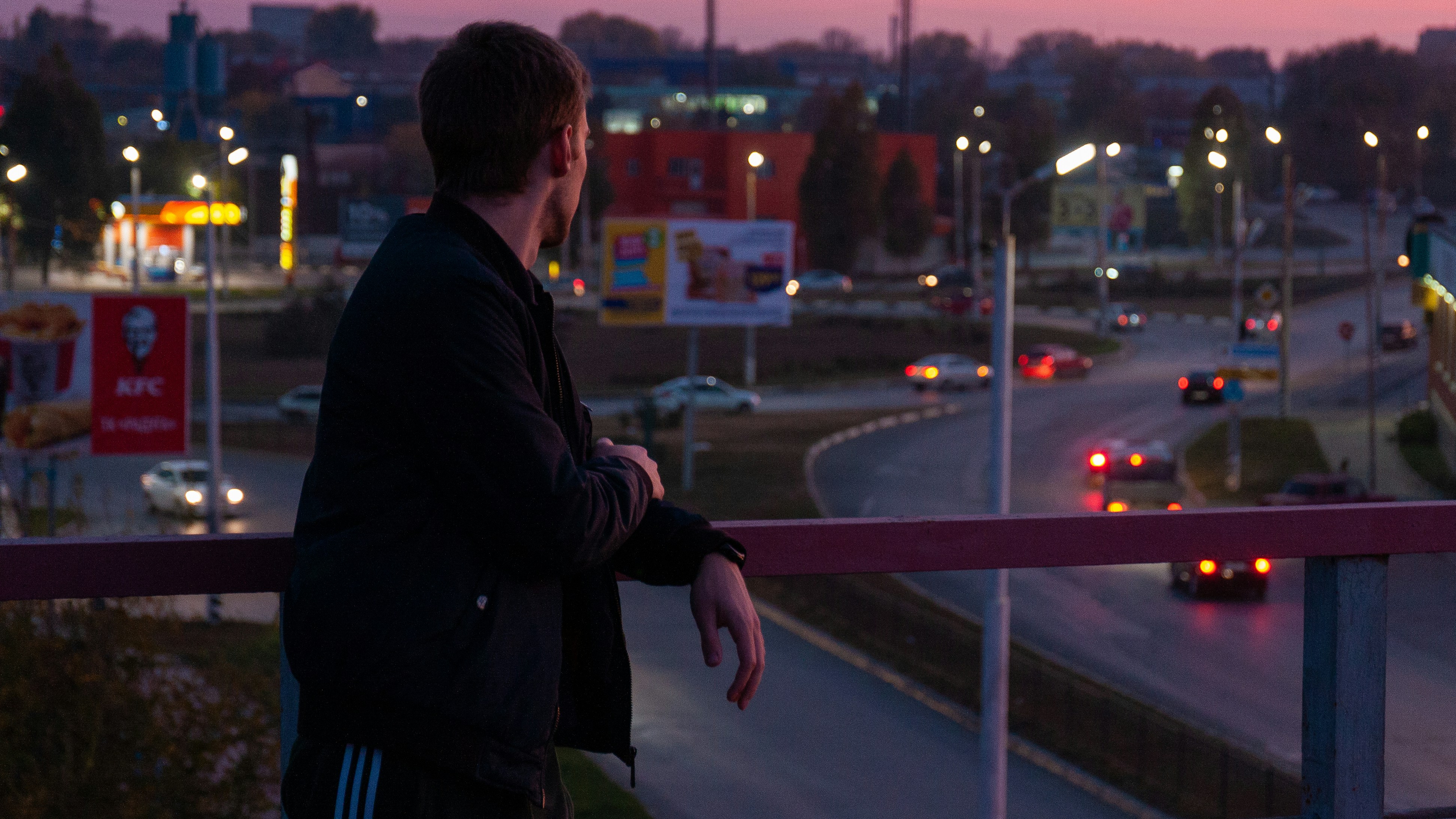 Person gazing over a city street at dusk with vibrant sky and streetlights illuminating the scene.