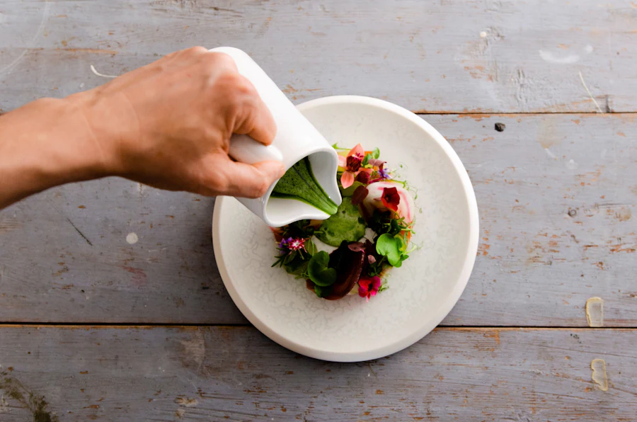 A hand tips a small white pitcher over a bowl of colorful salad greens on a weathered wooden table. Fresh vegetables and flowers add bright color to the minimalist plate.