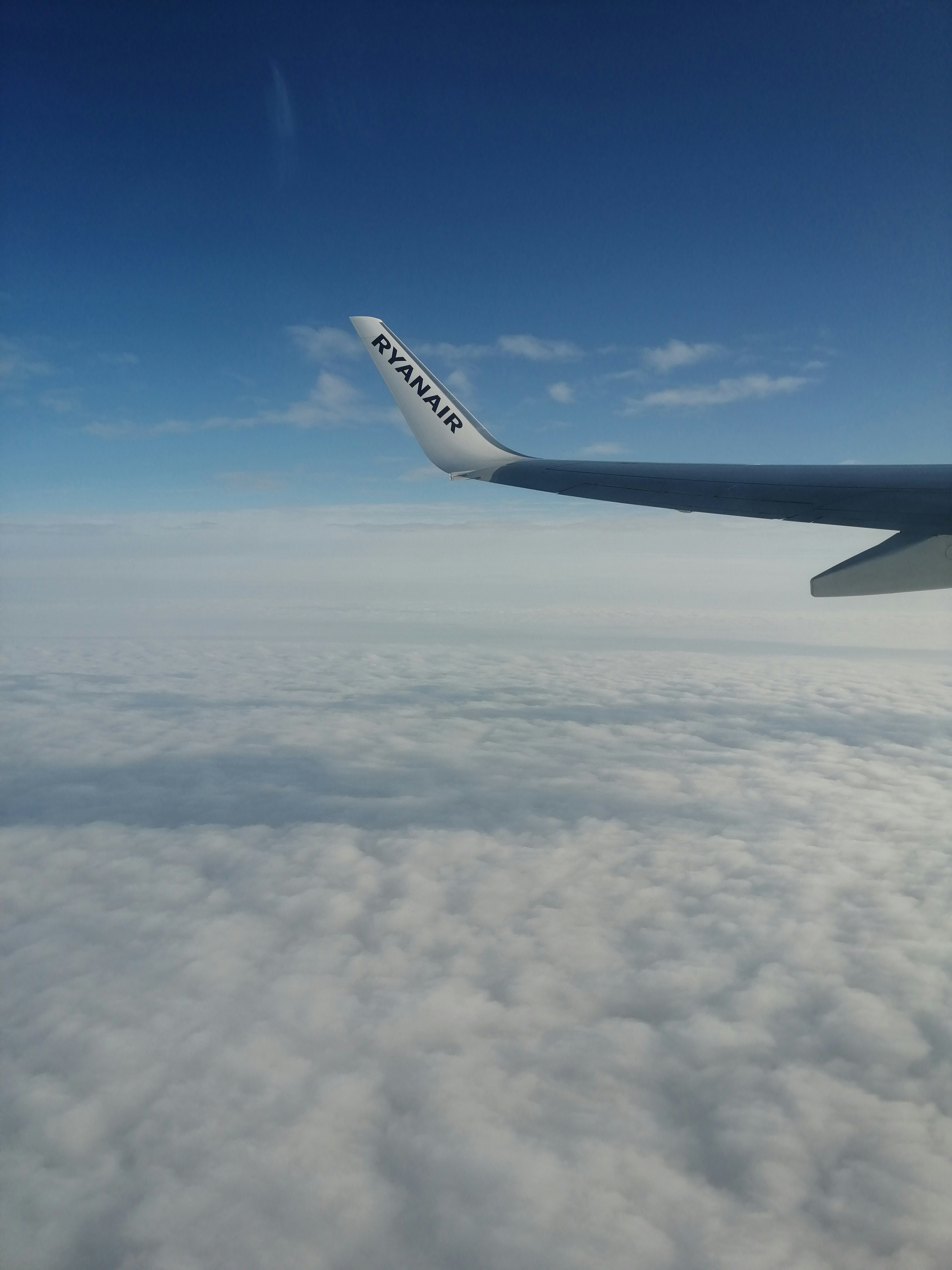 Airplane wing cutting through a sea of clouds under a bright blue sky. The scene captures the essence of travel and adventure.
