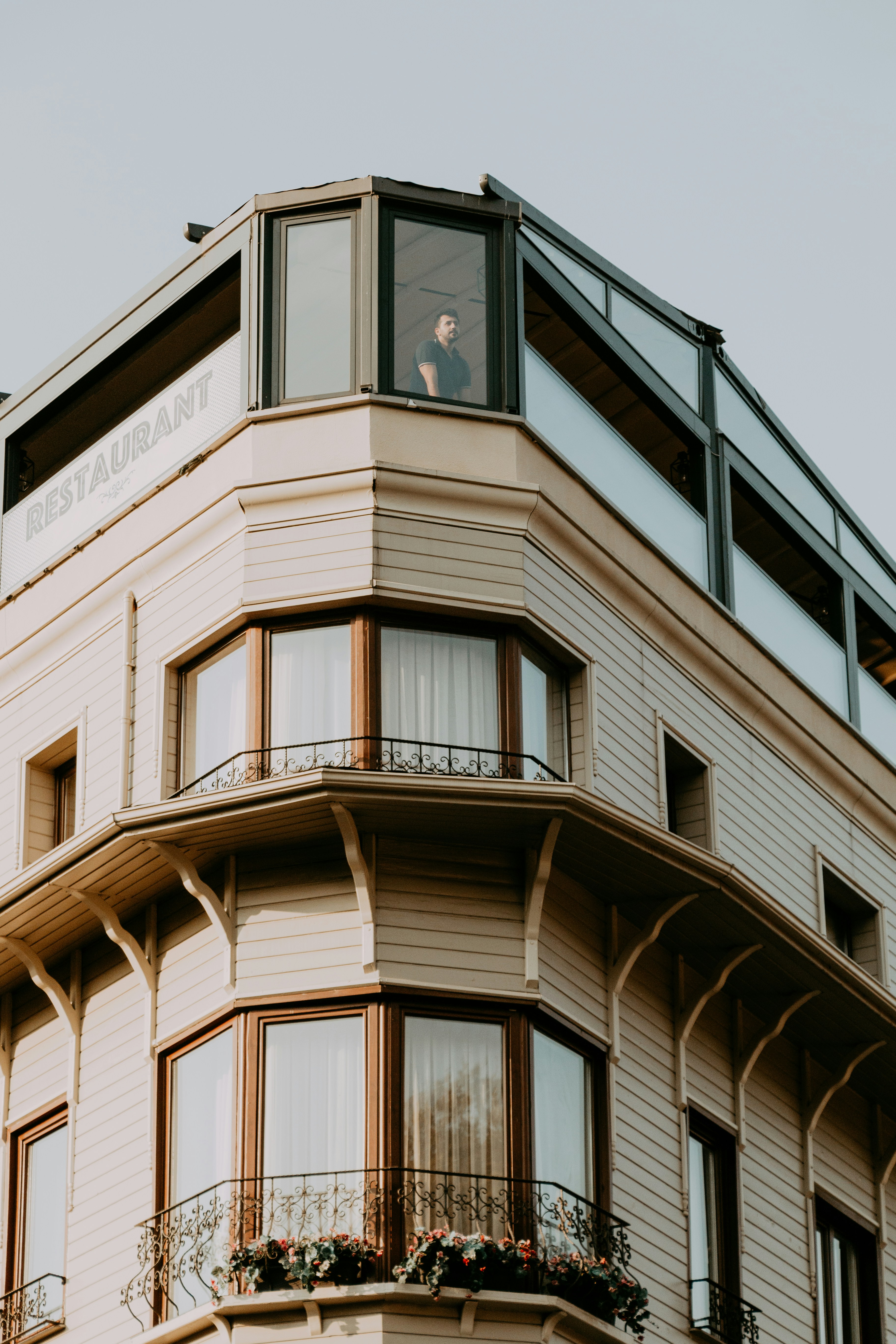 A person gazes out from a restaurant balcony, framed by the elegant architecture of the building below. The design features intricate details and a warm color palette.