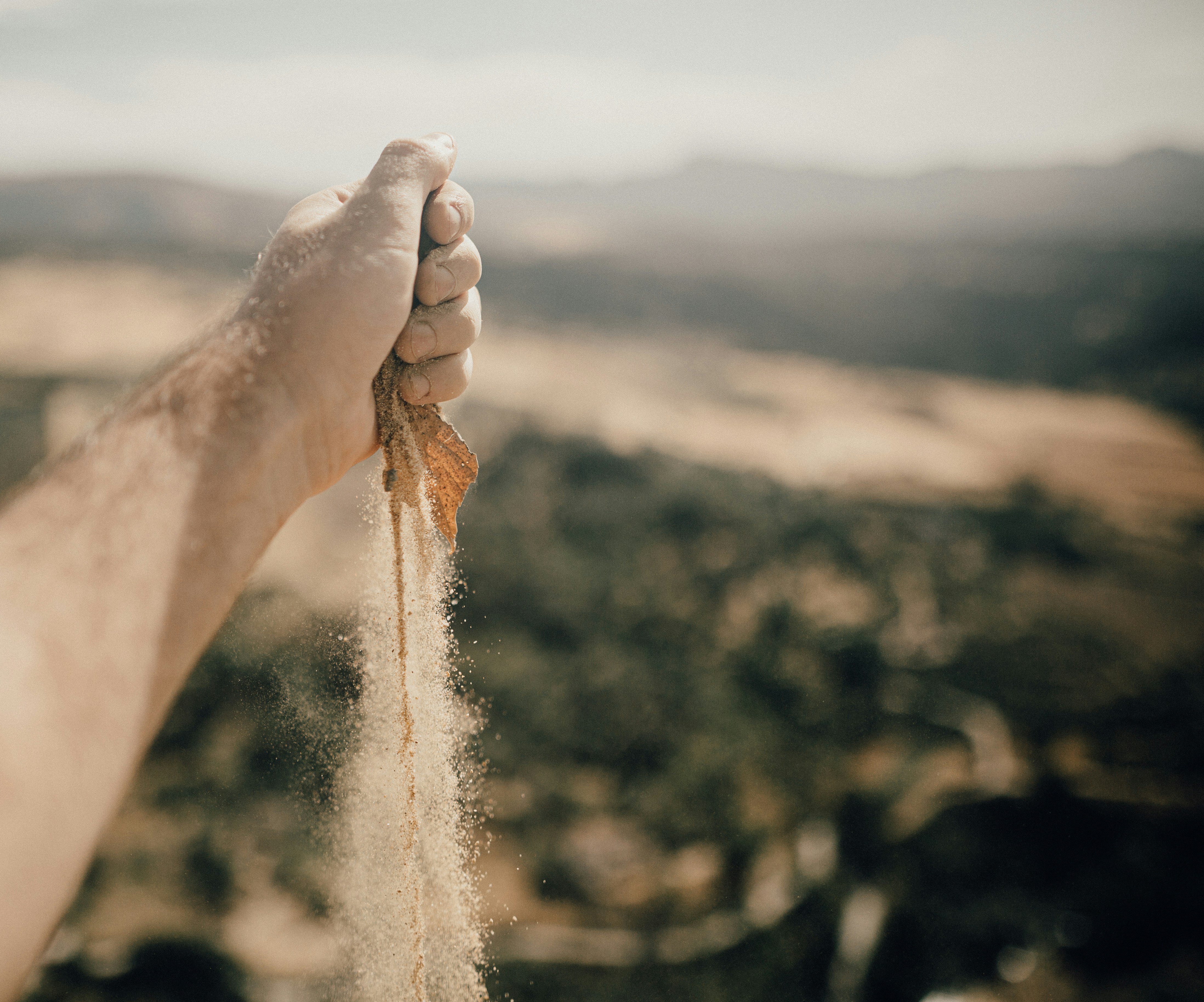 A hand releasing sand against a blurred landscape, symbolizing the passage of time and the transient nature of life.