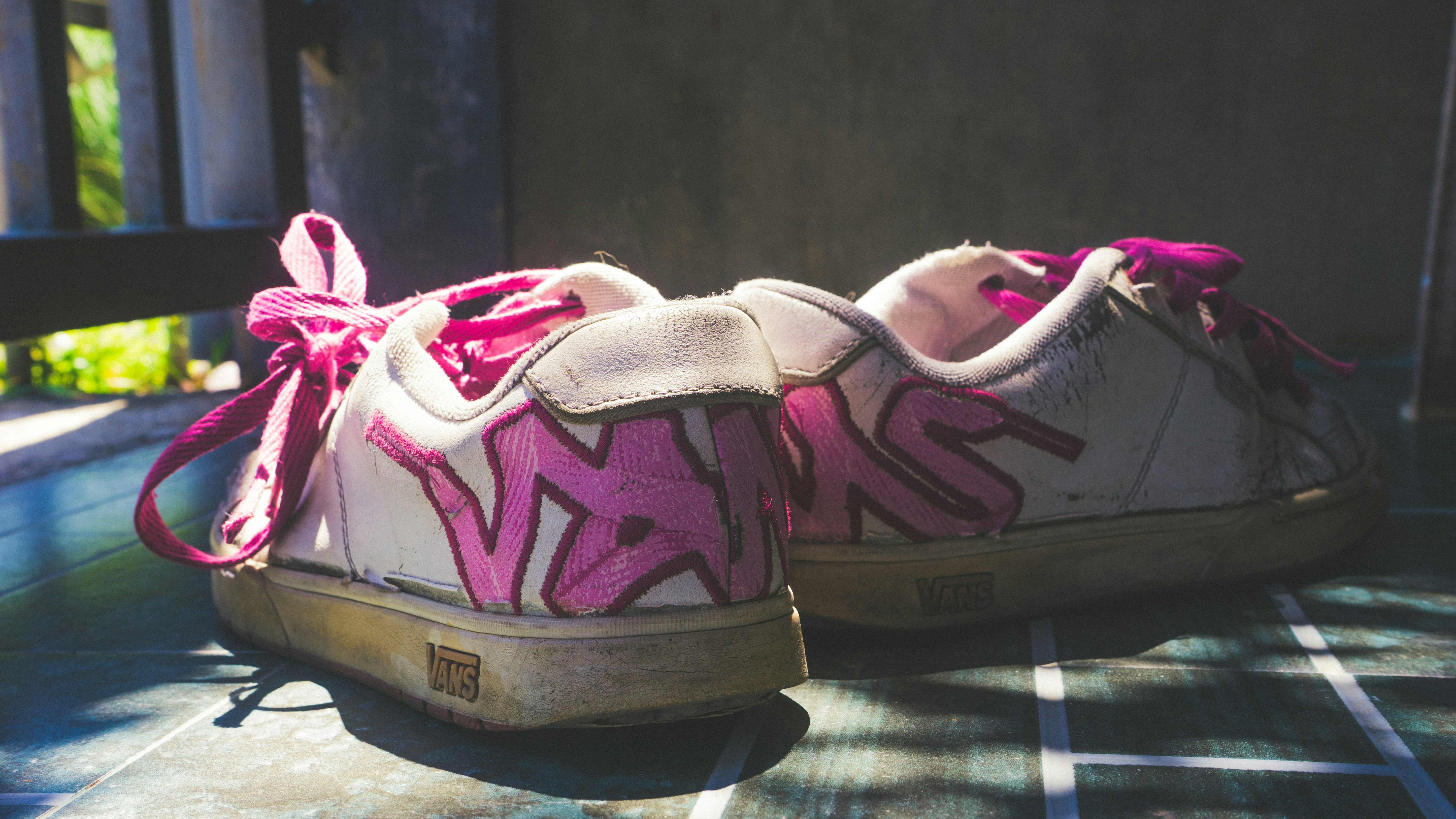 Worn white sneakers adorned with pink graffiti sit on a tiled surface, capturing a moment of urban life. The sunlight highlights their unique character.